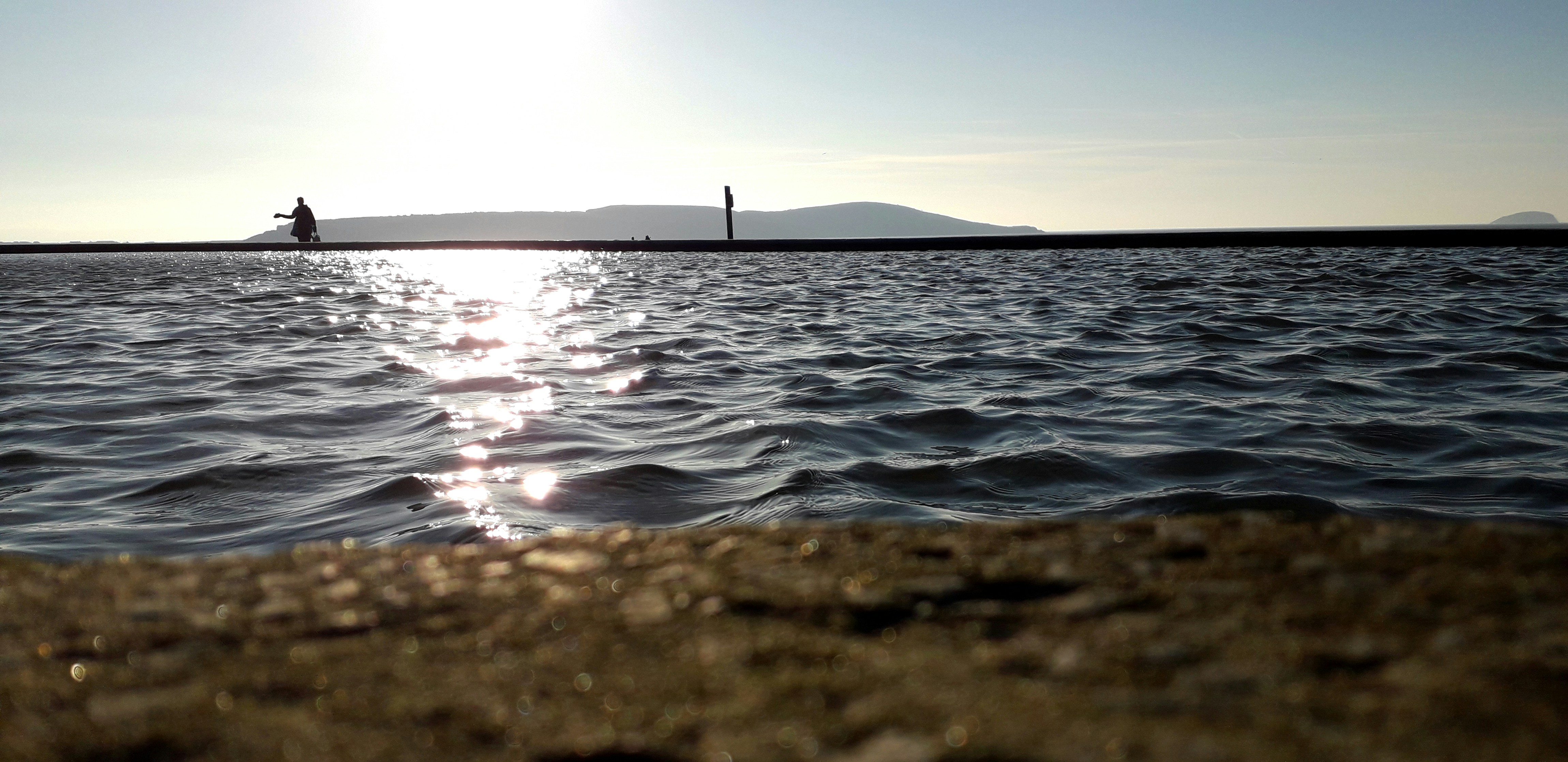 Silhouette of a person standing by the sea as sunlight reflects off gentle waves with distant hills visible.
