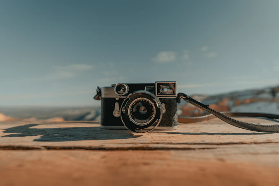 A vintage film camera sits on a weathered wooden surface with a blurred landscape and blue sky behind it. The strap stretches off to the right, and the camera casts a long shadow in warm light.