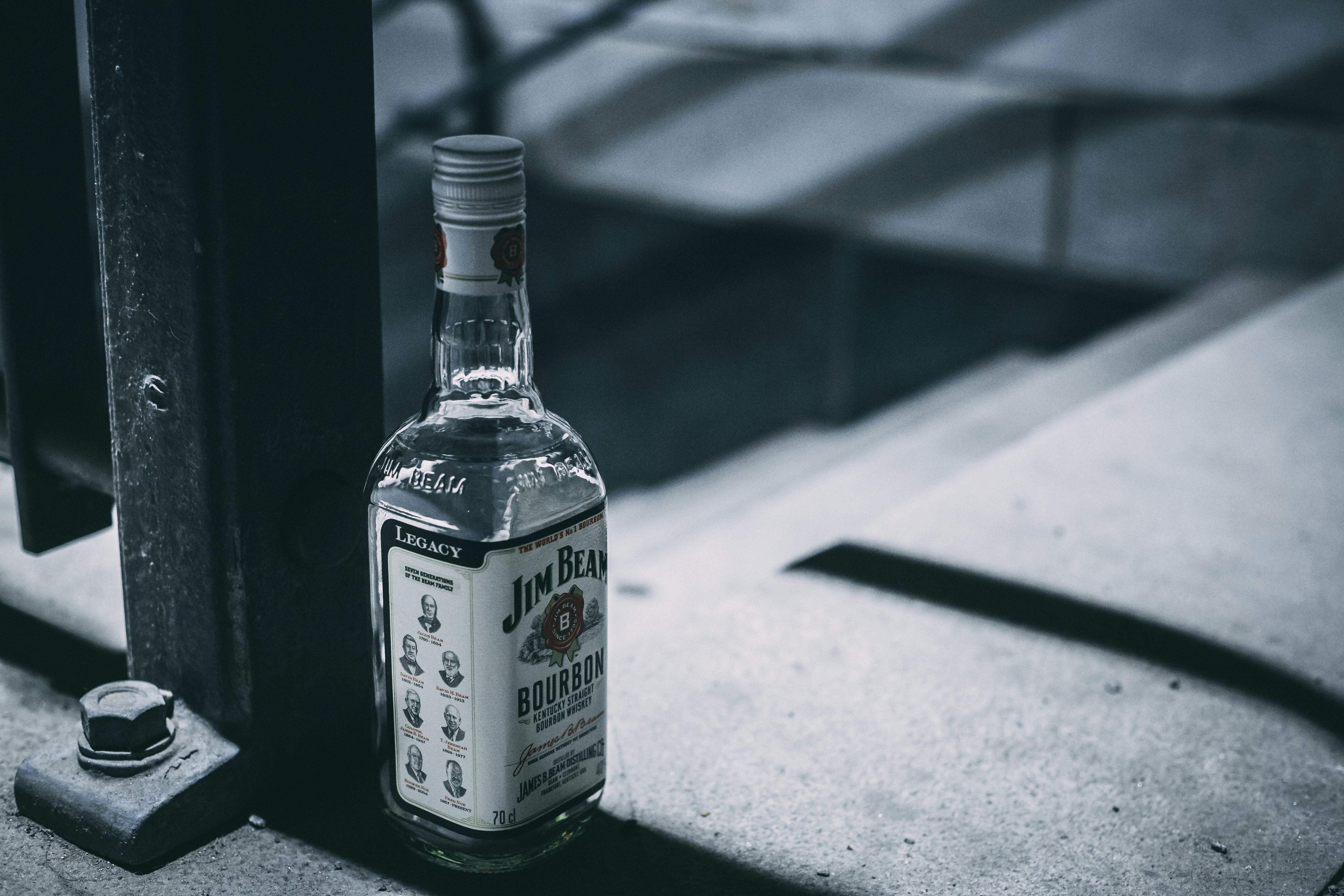 A bottle of Jim Beam bourbon rests against a metal railing, casting shadows on the concrete steps. The monochromatic tones enhance the scene's nostalgic atmosphere.