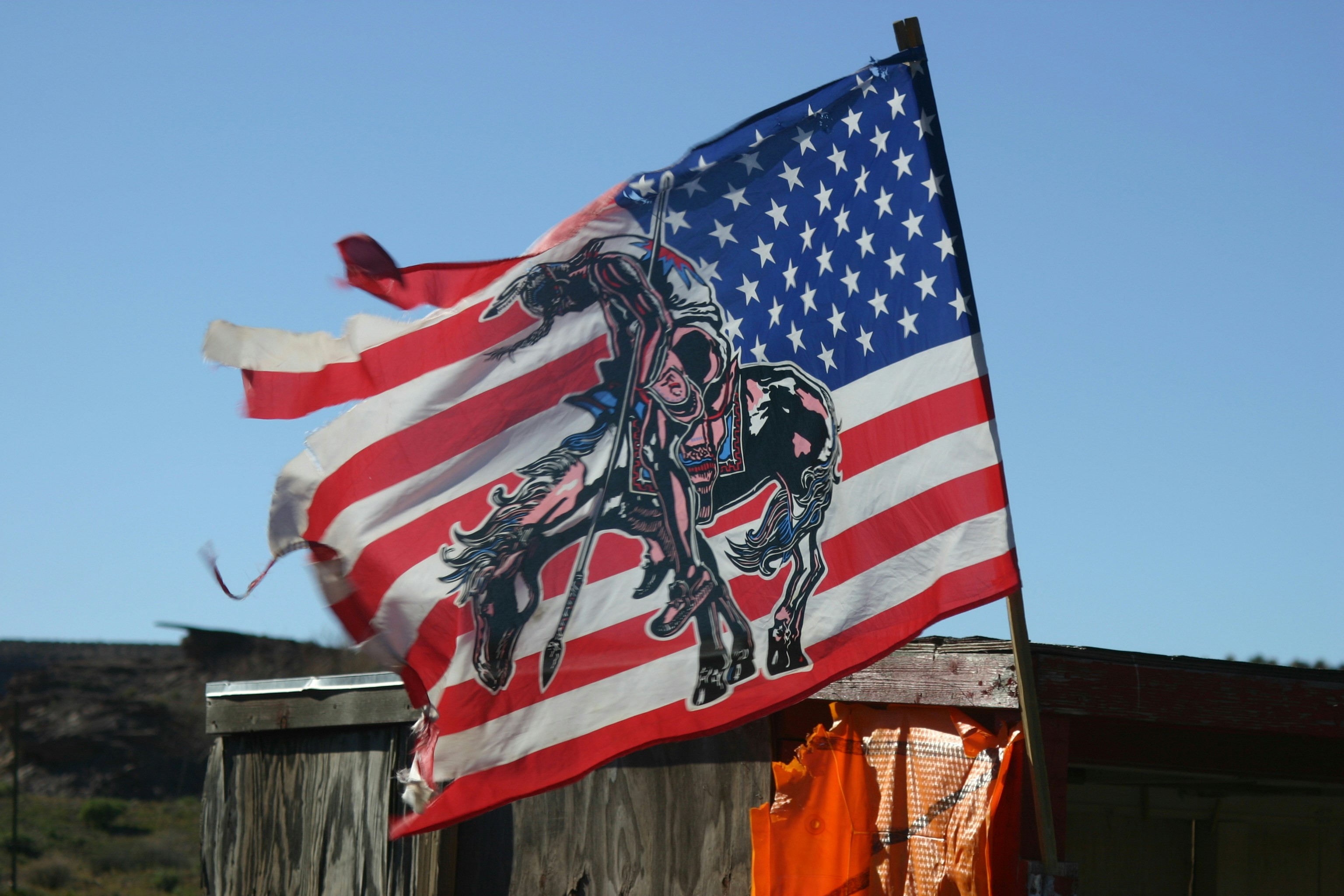 A weathered flag featuring a cowboy silhouette blends with the American flag, fluttering against a clear blue sky.