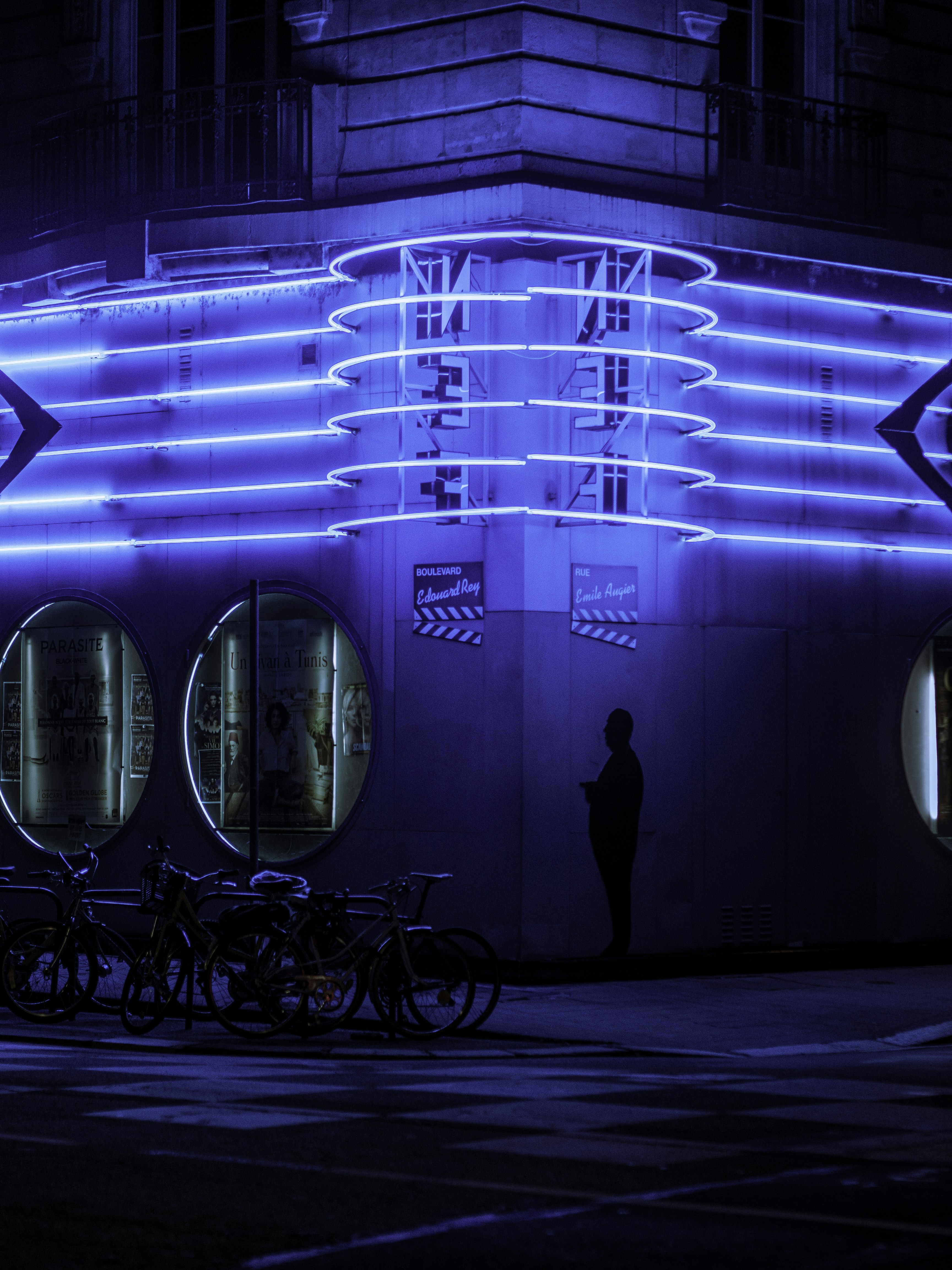 A vibrant neon-lit corner of a city at night, featuring circular windows and a solitary figure standing against the wall. The electric blue glow creates a dramatic atmosphere.