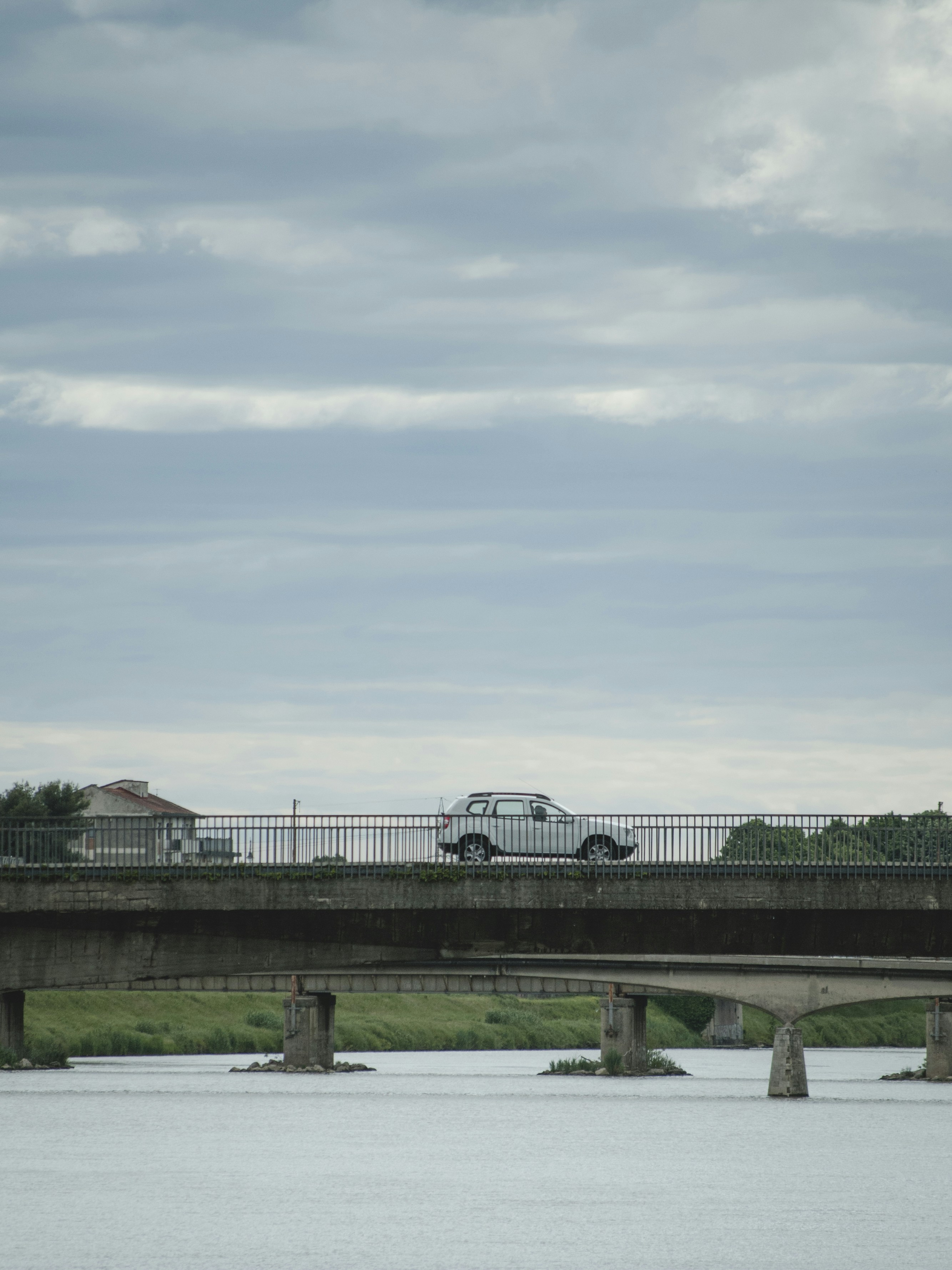 A solitary vehicle traverses a bridge over a serene river, framed by a cloudy sky. The scene evokes a sense of calm and contemplation.