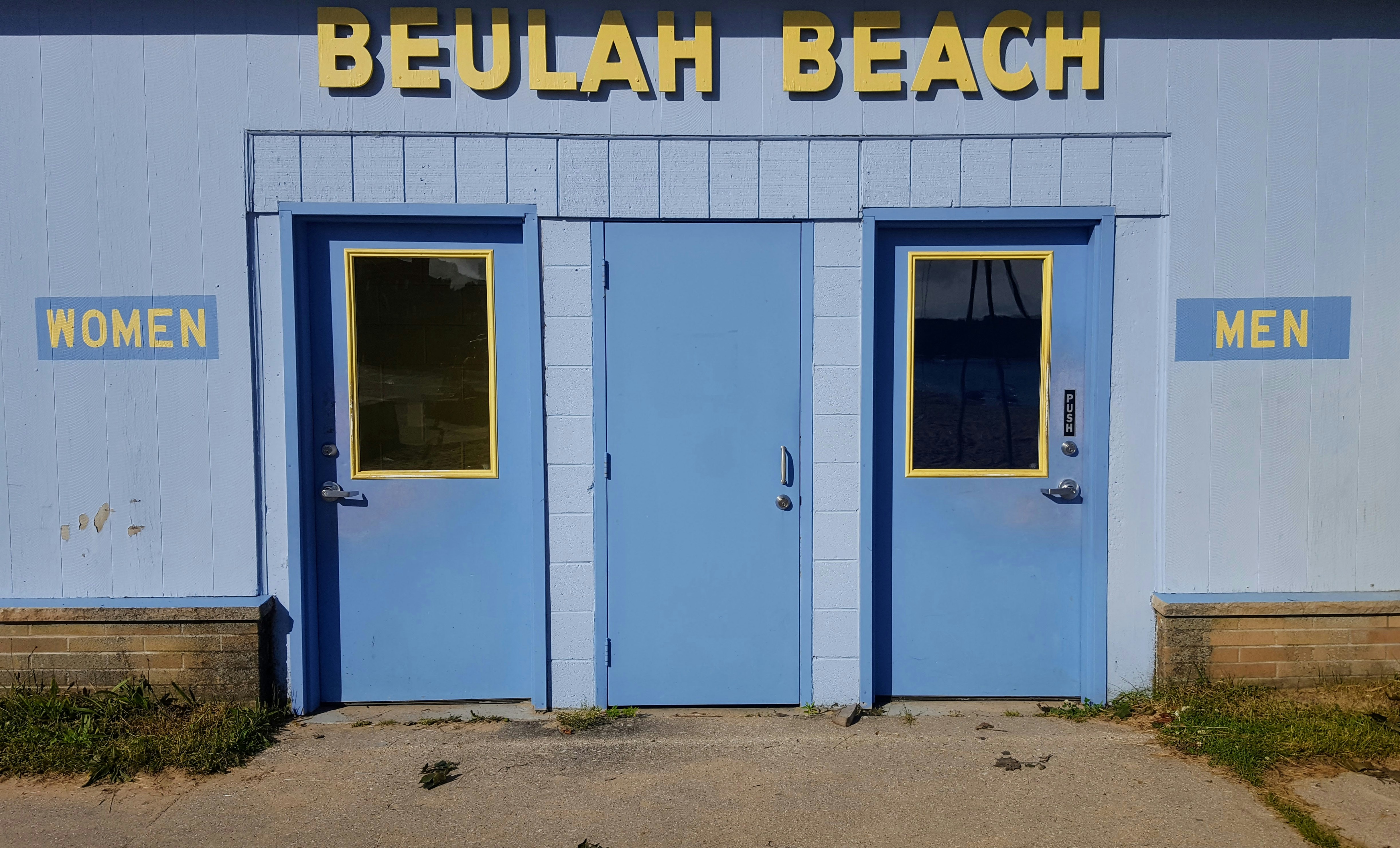Powder-blue beach facility facade with two doors and matching windows, labeled 'WOMEN' and 'MEN' beside the central entry under a 'BEULAH BEACH' sign.