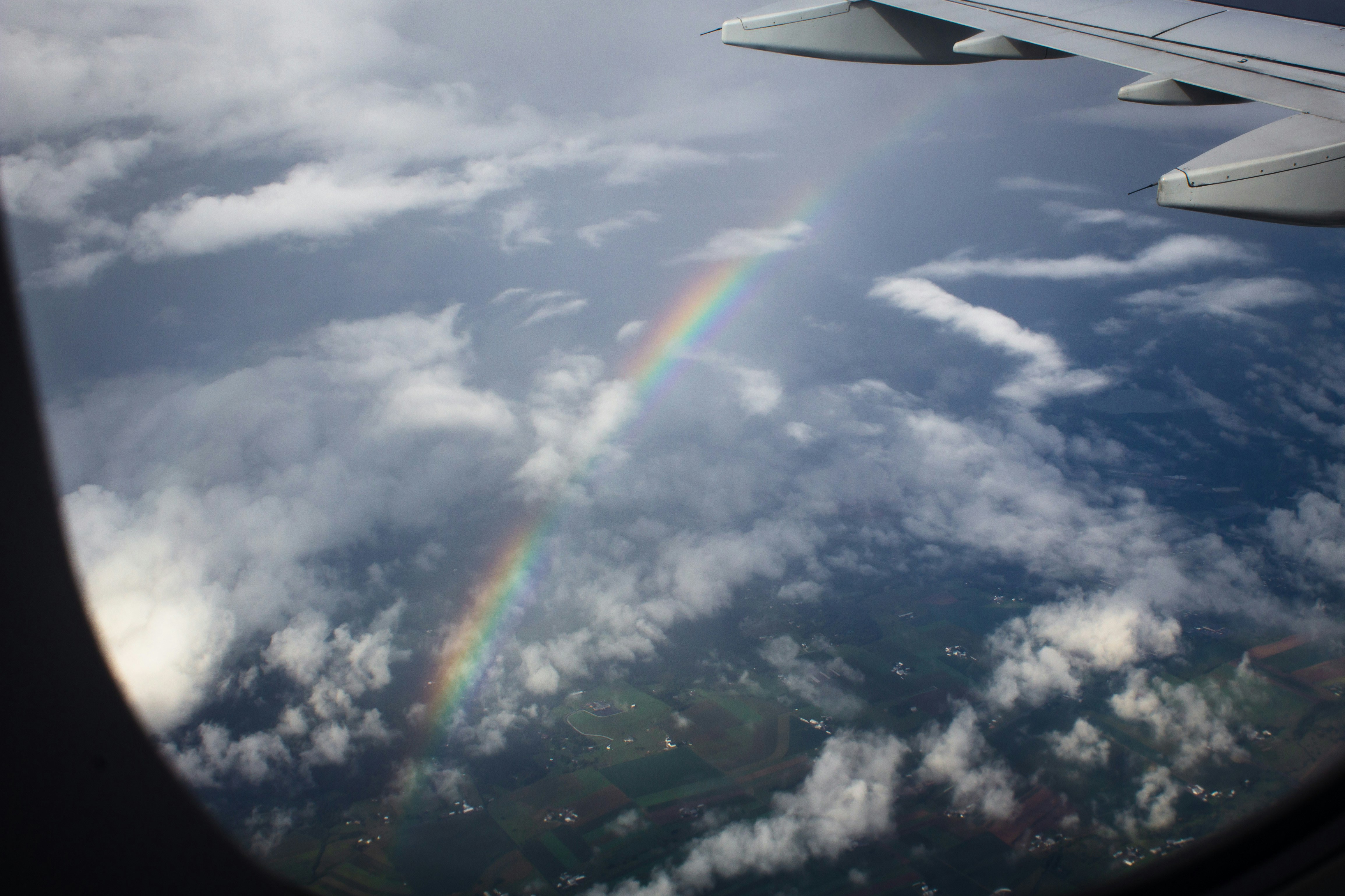 Rainbow arching over lush landscapes viewed from an airplane window, framed by clouds and wing. 