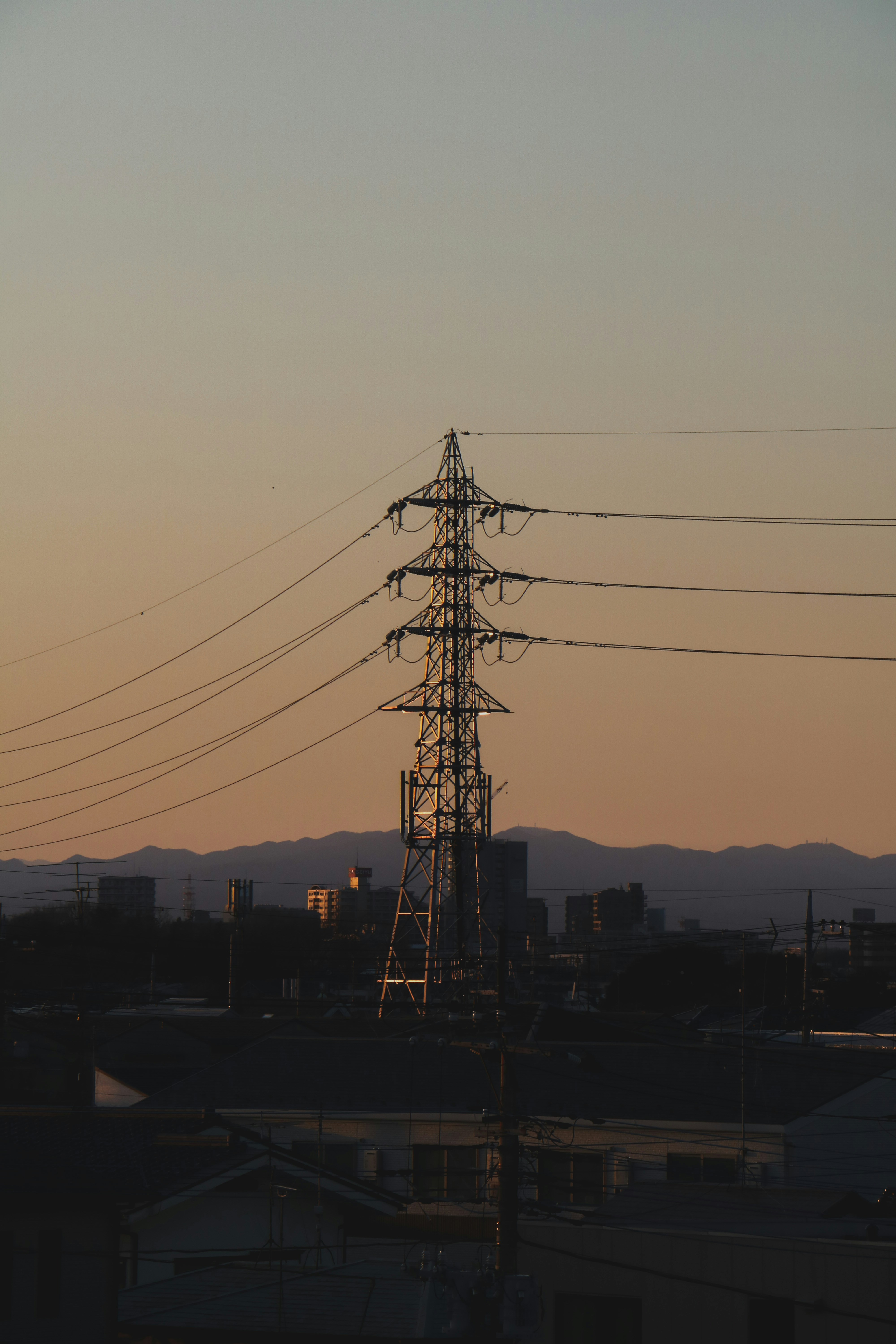 Silhouetted power tower against a gradient sunset, with distant mountains and urban landscape in the background.