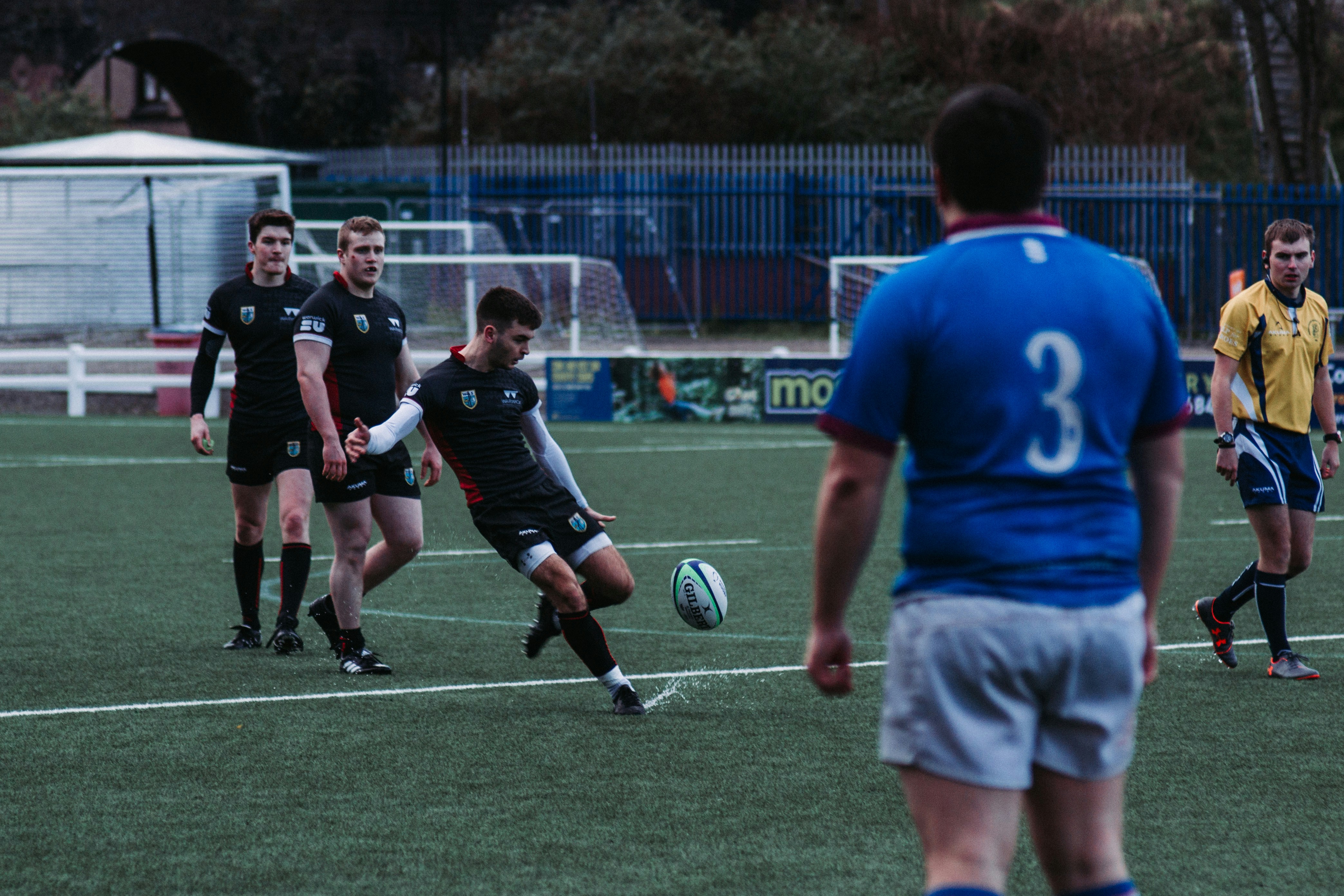 Rugby players poised on the field, with one player preparing to kick the ball while teammates and opponents observe the play.