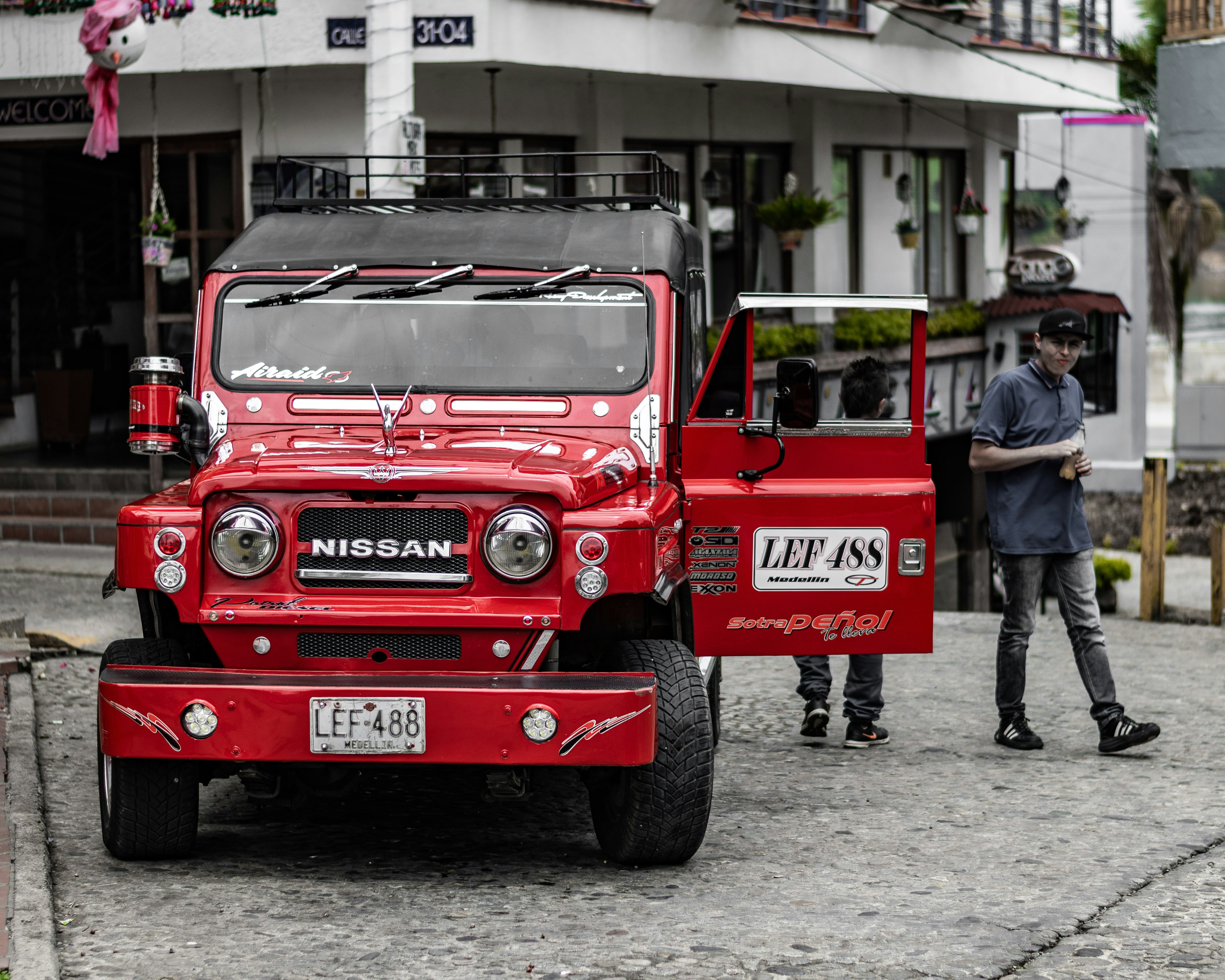 A vibrant red Nissan vehicle parked on a cobblestone street, with its door open and two individuals nearby engaged in conversation.
