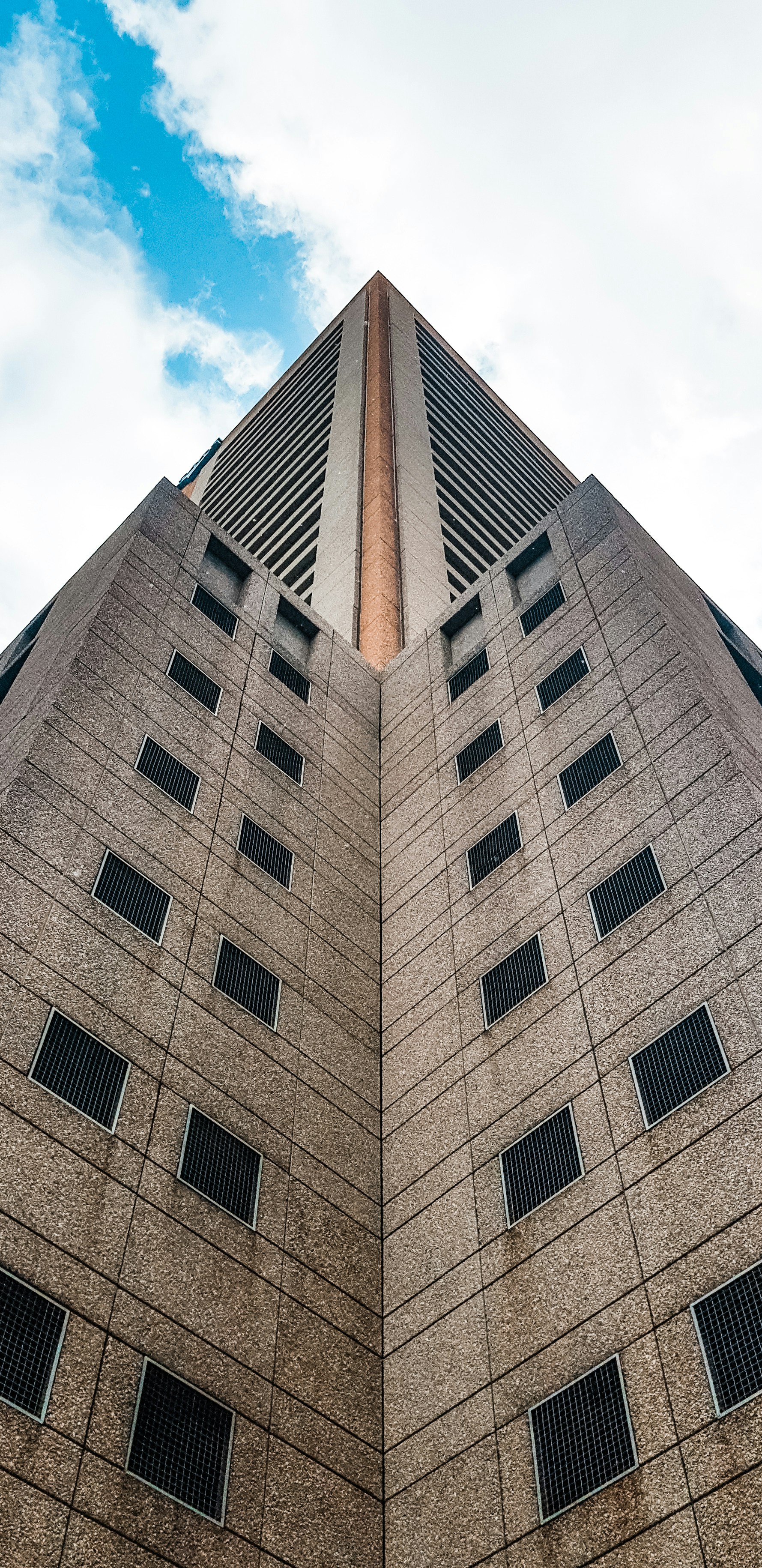 A towering building viewed from below, showcasing its textured facade and grid-patterned windows against a bright sky.