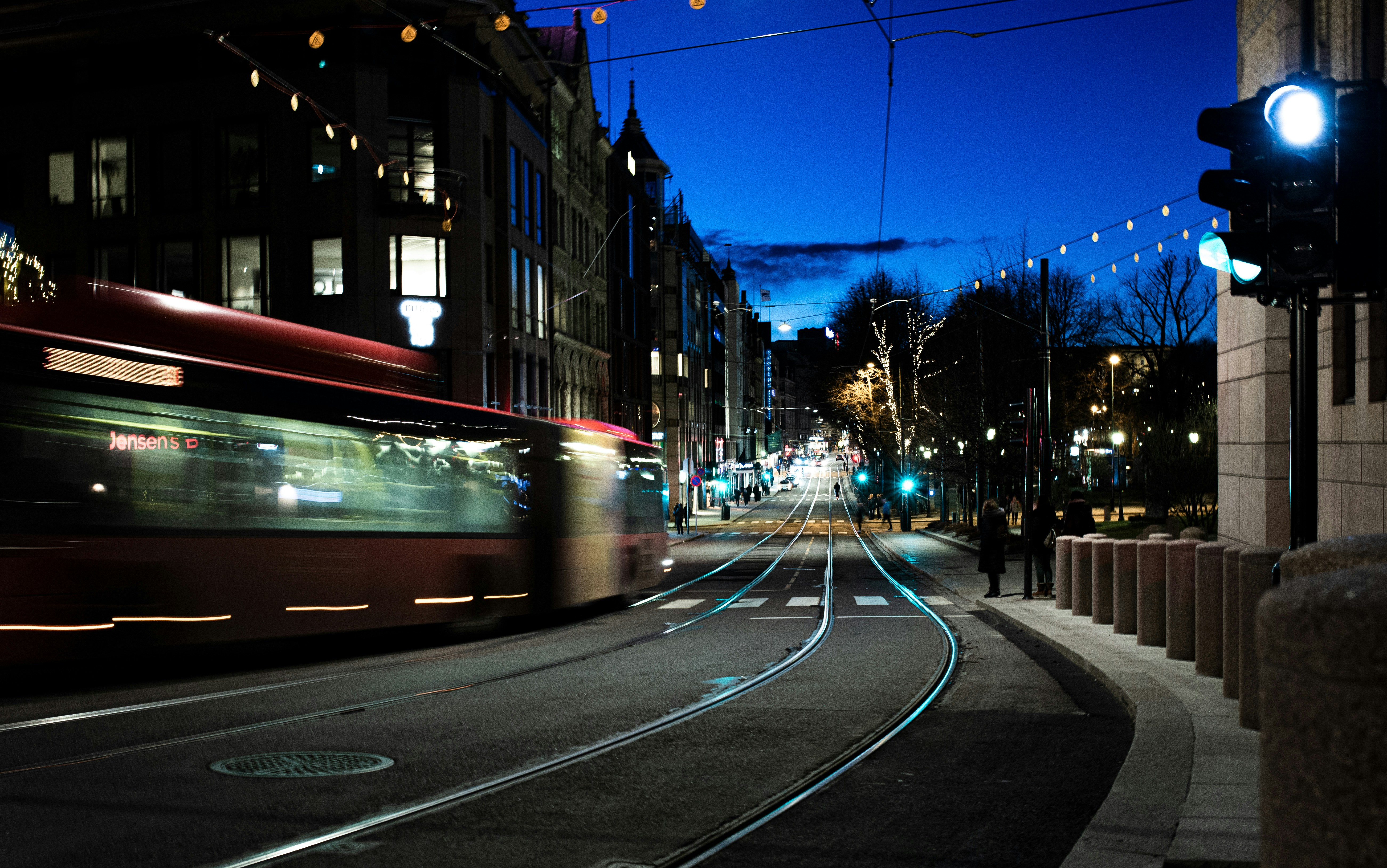 Blurred tram speeding through a city street at dusk with illuminated buildings and trees.