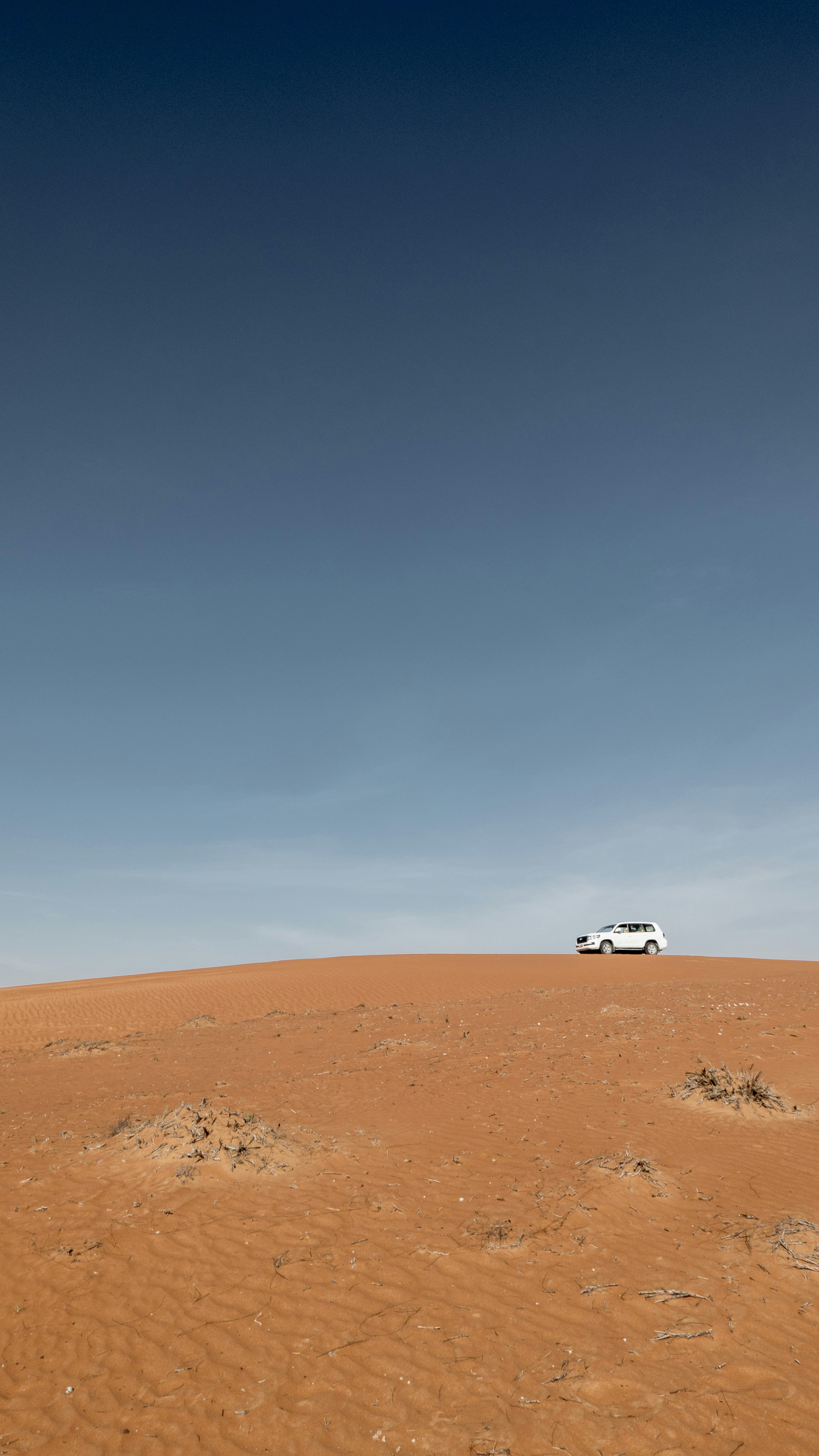 A white vehicle traversing a vast expanse of orange sand dunes under a clear blue sky.