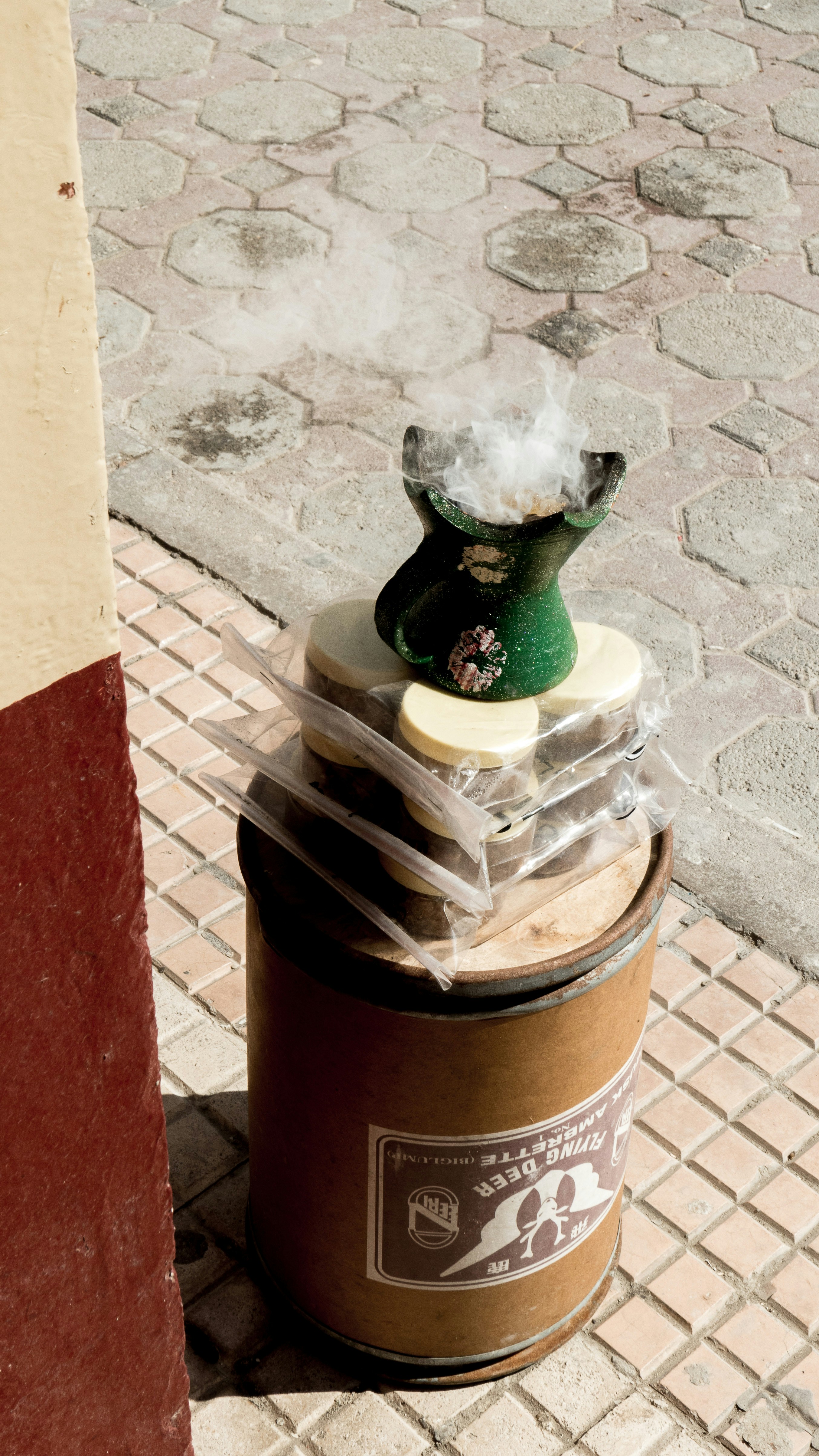 A vibrant green incense burner emits fragrant smoke, perched atop a wooden barrel beside a cobblestone street. The scene captures the essence of local culture and culinary tradition.