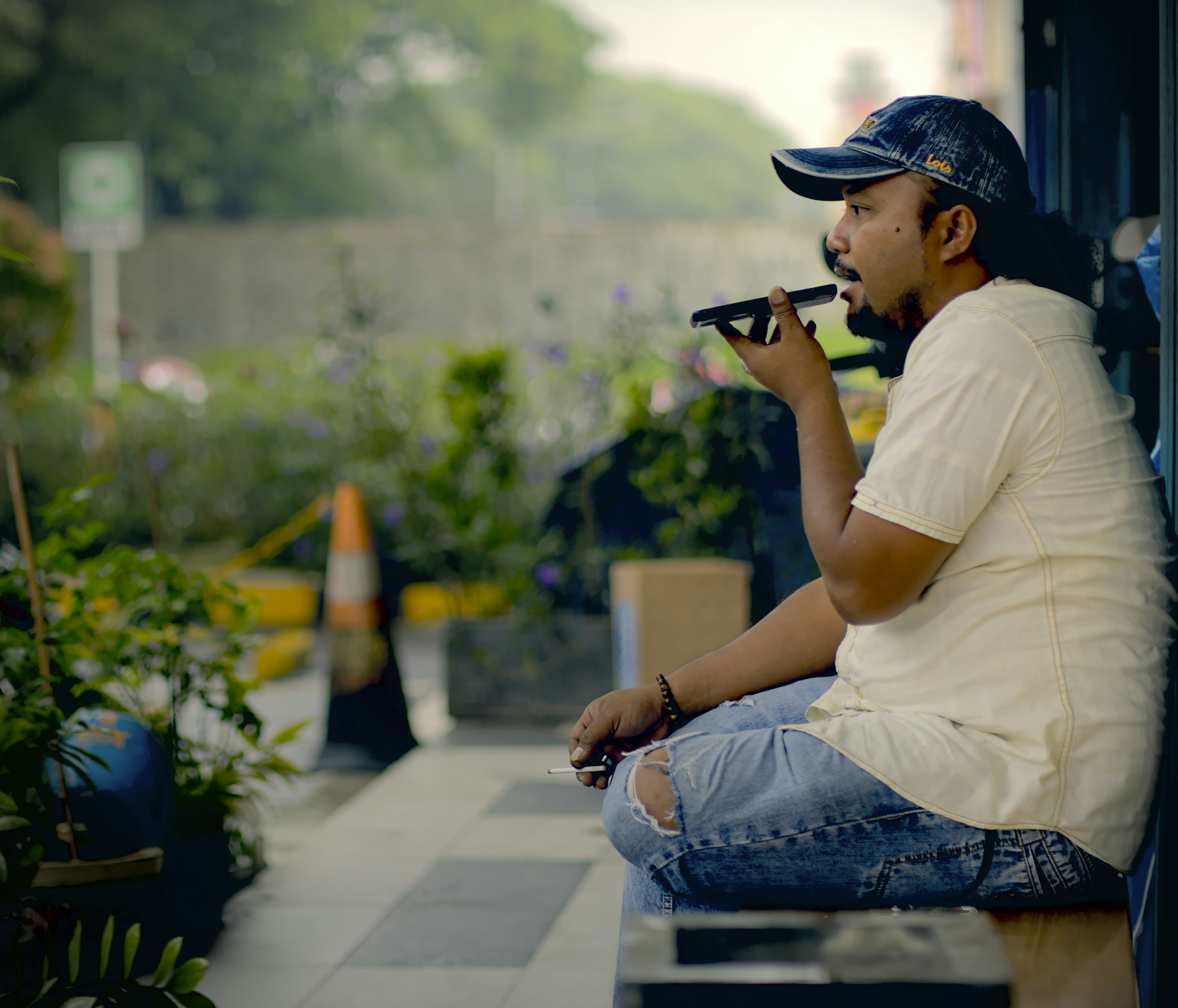 Man seated on a sidewalk, engaged in thought while smoking, surrounded by greenery and urban elements.