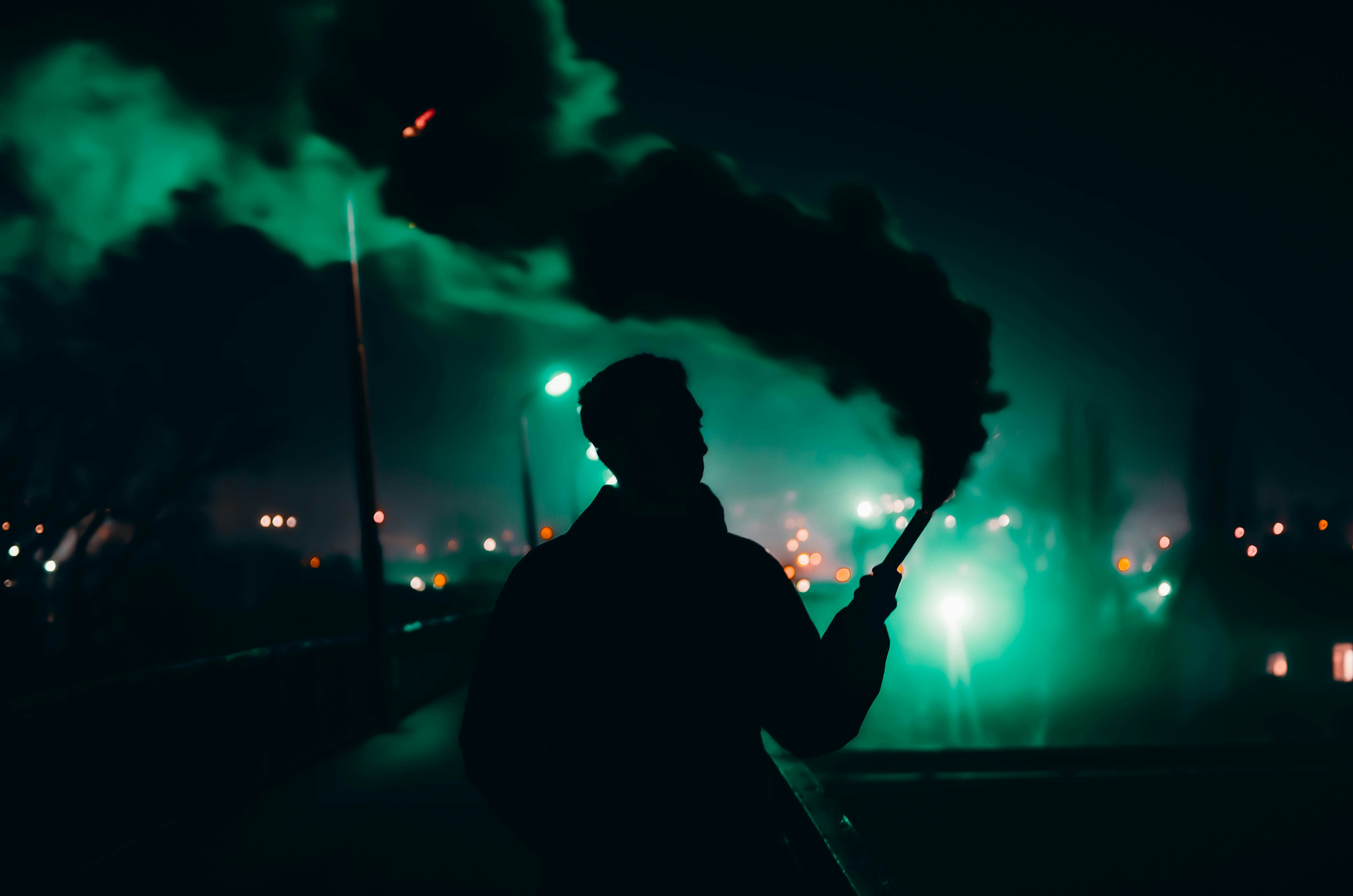 Emerald Veil over the Night: Silhouette grips a green flare amid smoke and distant city lights