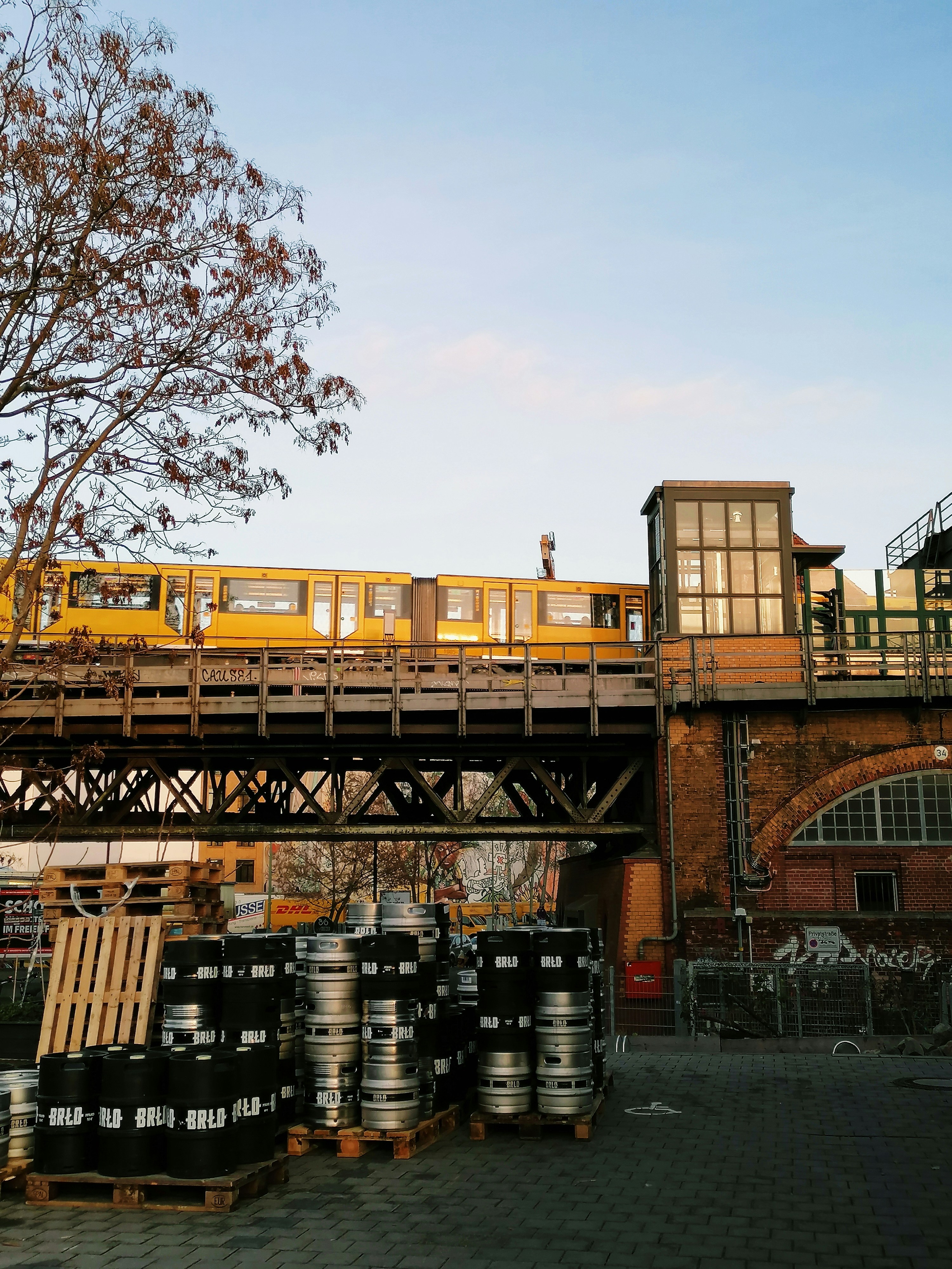 Yellow passenger train crossing an elevated steel bridge above a yard of stacked tires, with brick buildings and a clear blue sky.