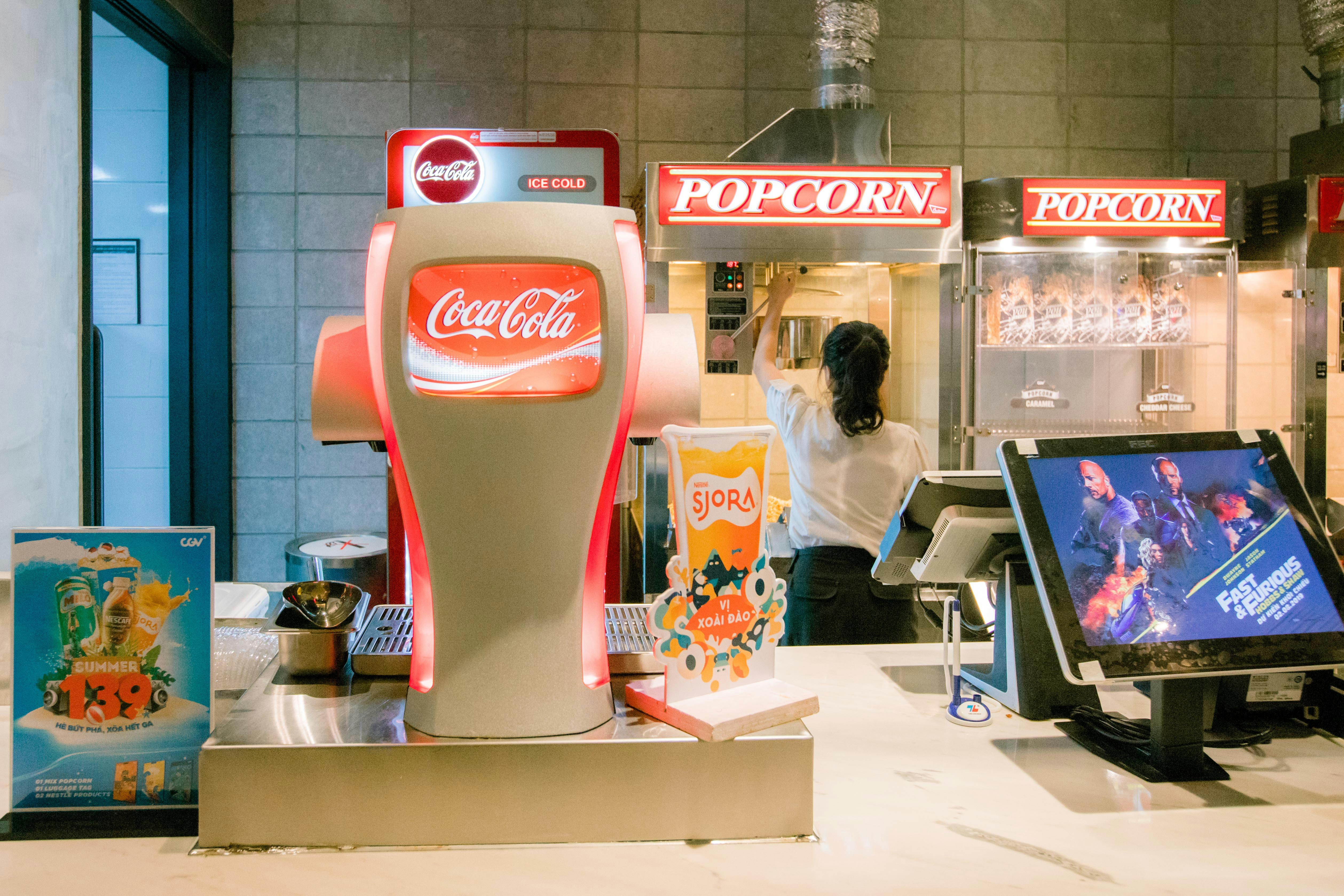 Coca-Cola dispenser at a cinema concession stand with popcorn and digital display.
