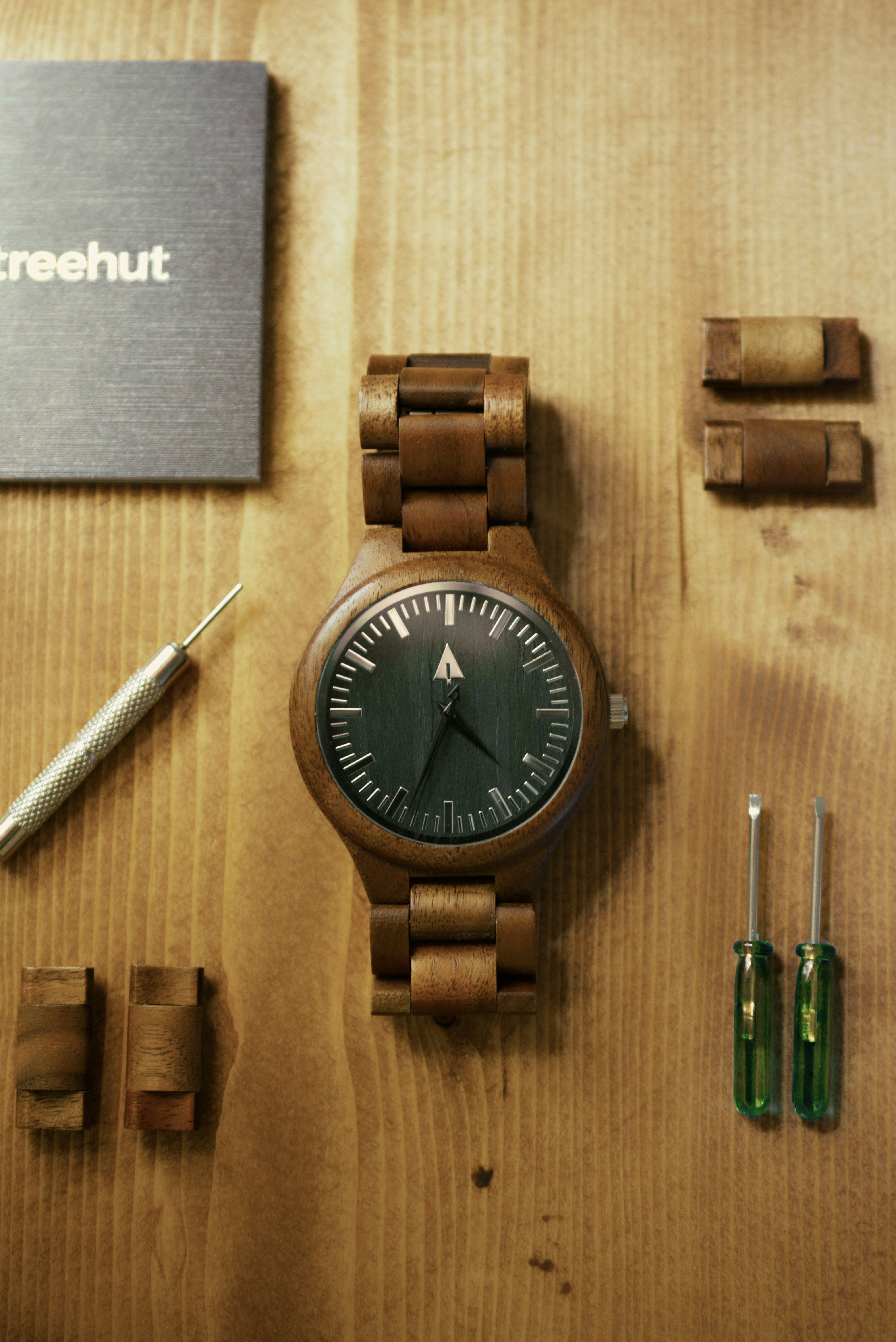 A wooden watch rests on a wooden surface, accompanied by tools and a branded card, showcasing the blend of nature and craftsmanship.