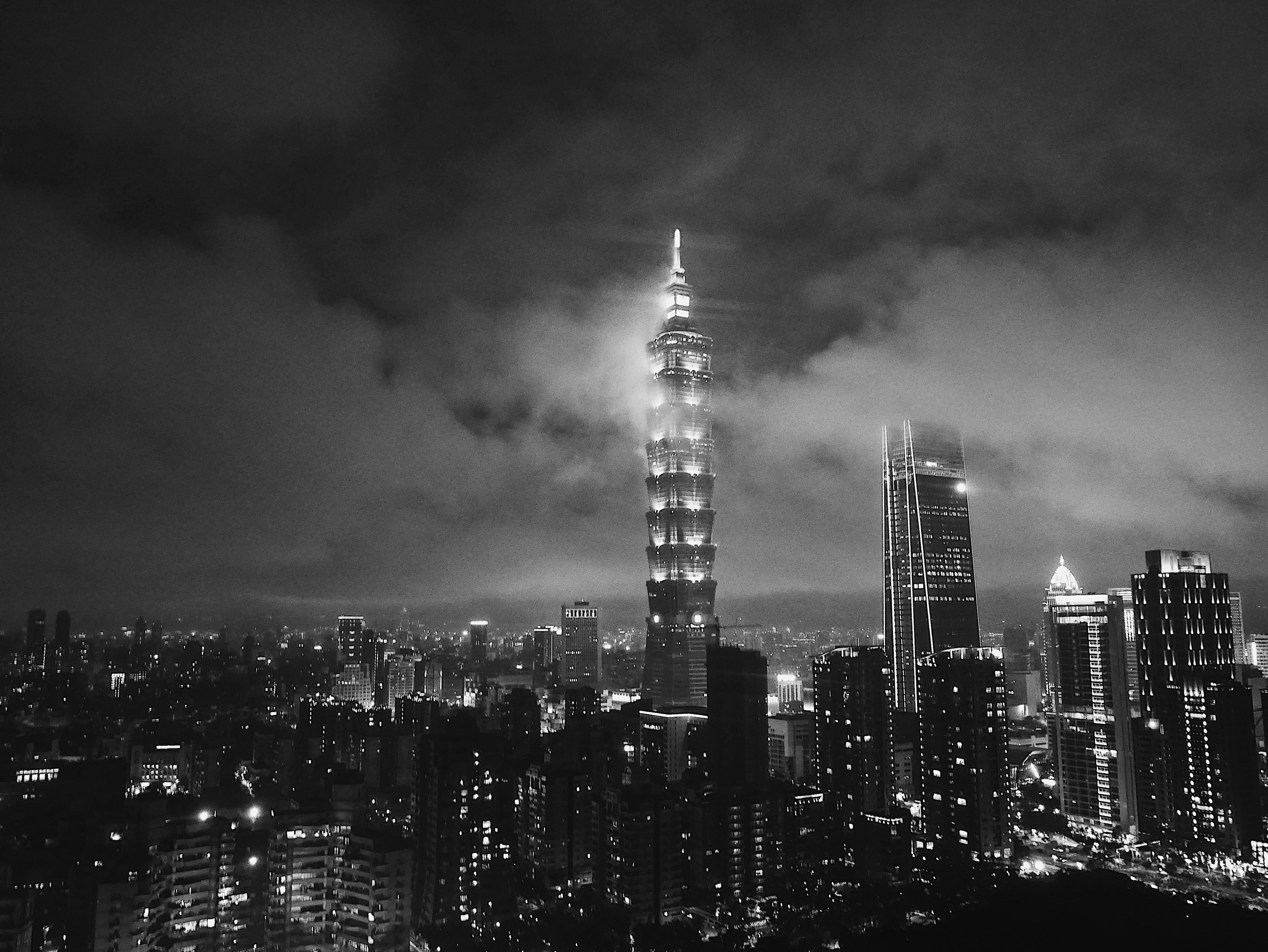 Taipei 101 piercing through low-hanging clouds, surrounded by a sea of illuminated skyscrapers at night.