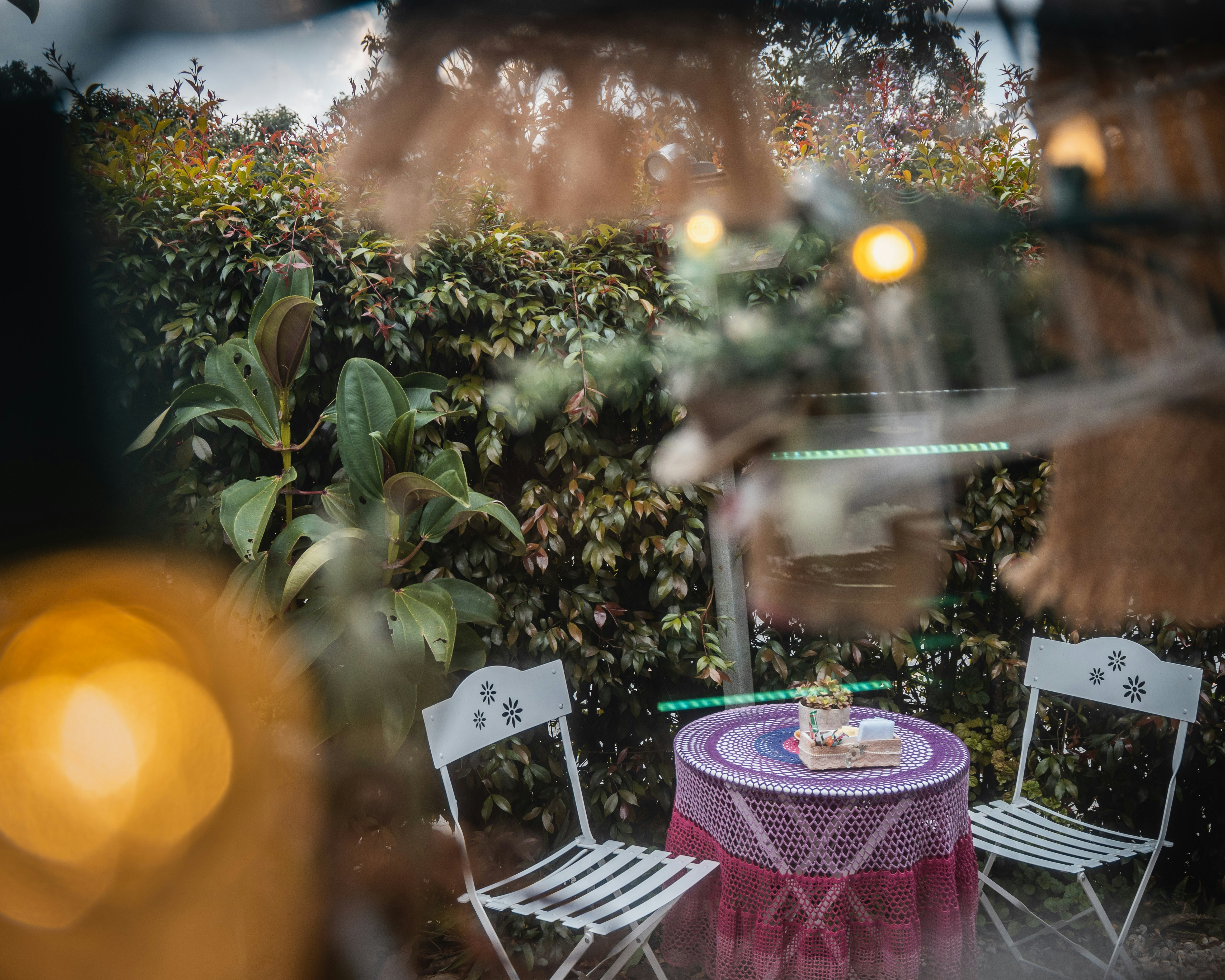 Cozy outdoor seating arrangement with a round table adorned with a tablecloth, surrounded by lush greenery and soft lighting.