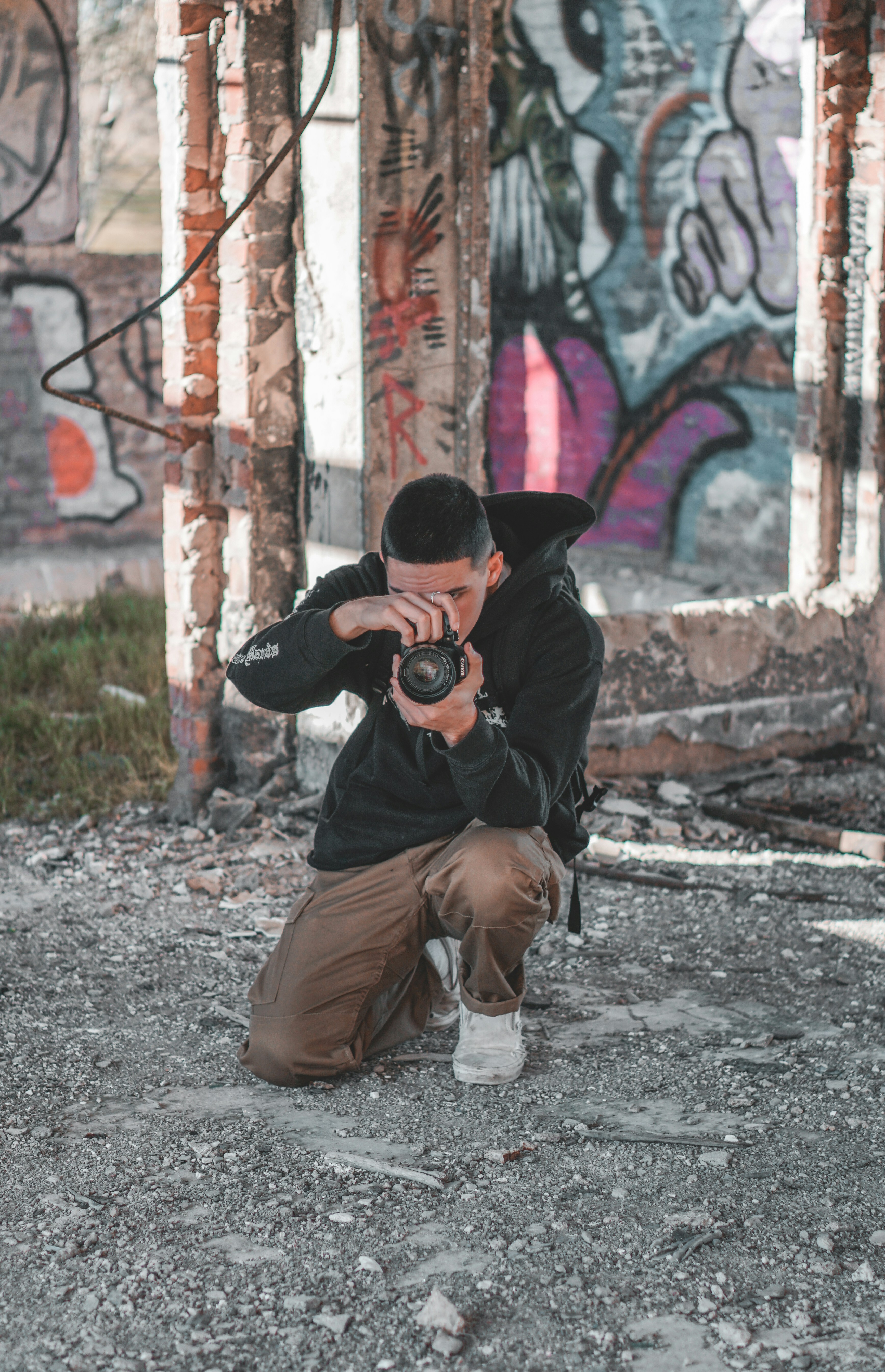 A person crouches in an abandoned building, focusing intently through a camera, surrounded by colorful graffiti and remnants of decay.