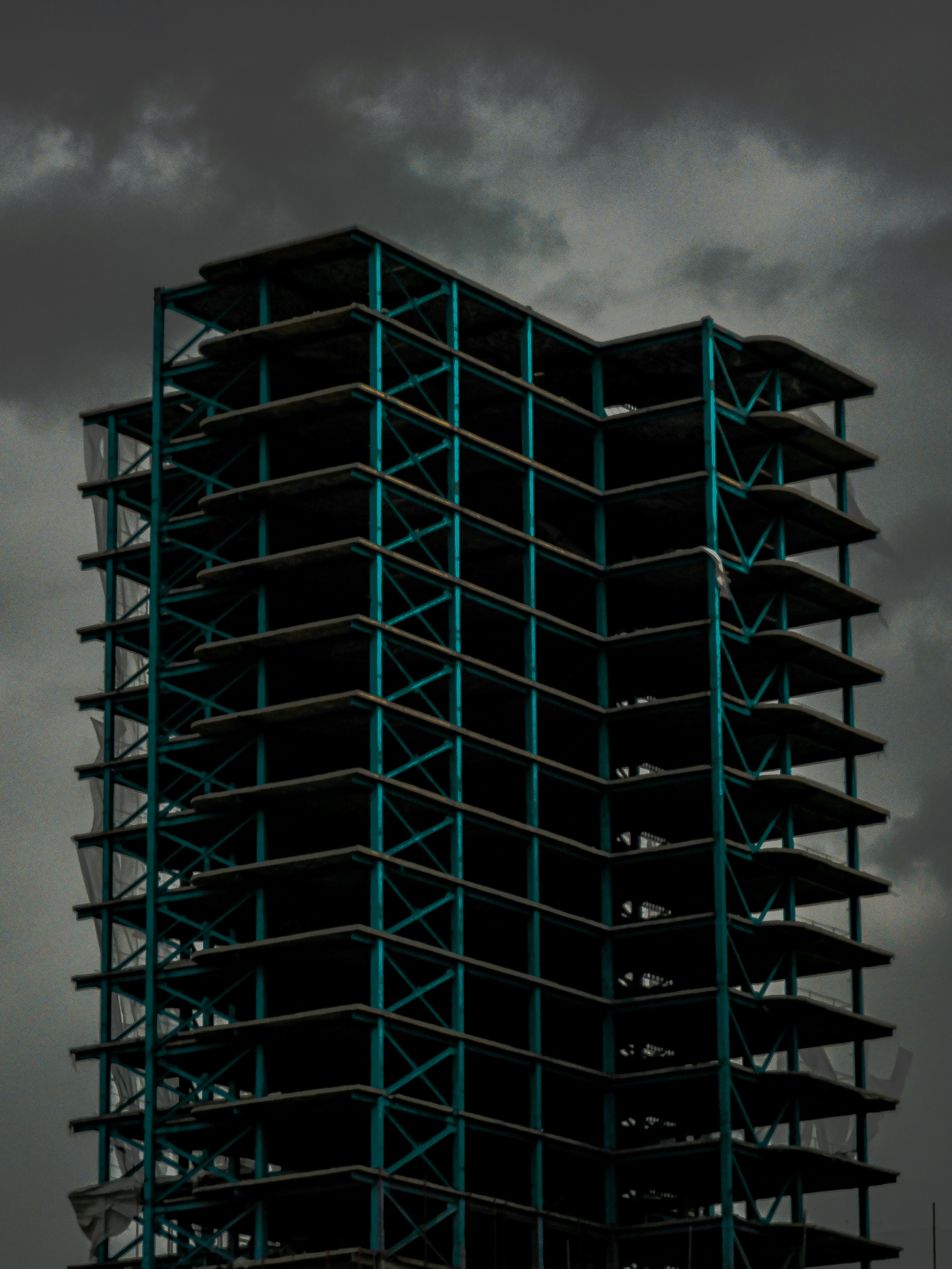 Unfinished high-rise skeleton with teal diagonal braces rises against dark, overcast skies. The stark steel framework dominates the frame, emphasizing construction and foreboding weather.