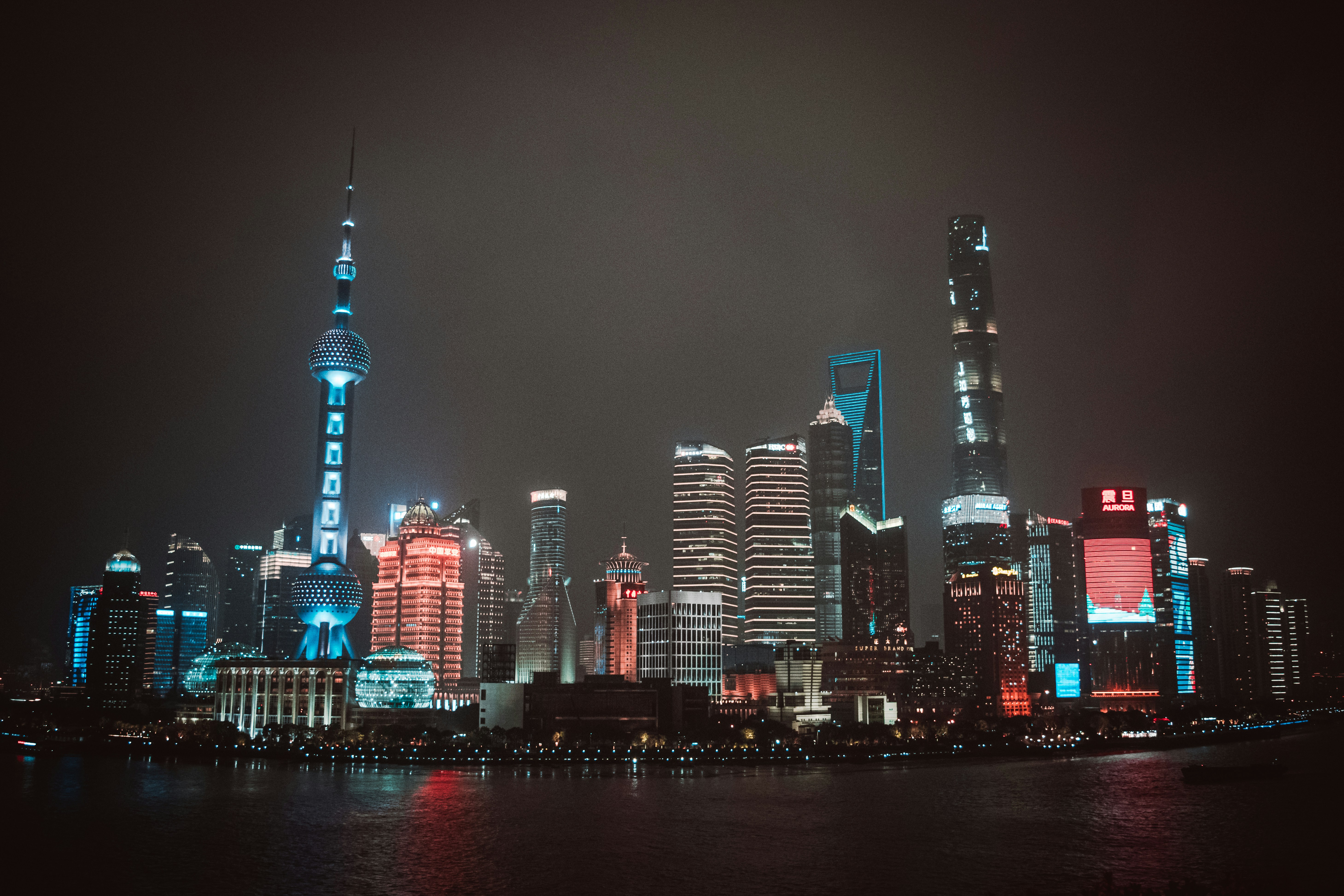 Illuminated skyline of Shanghai featuring the Oriental Pearl Tower and modern skyscrapers reflecting vibrant colors on the water.