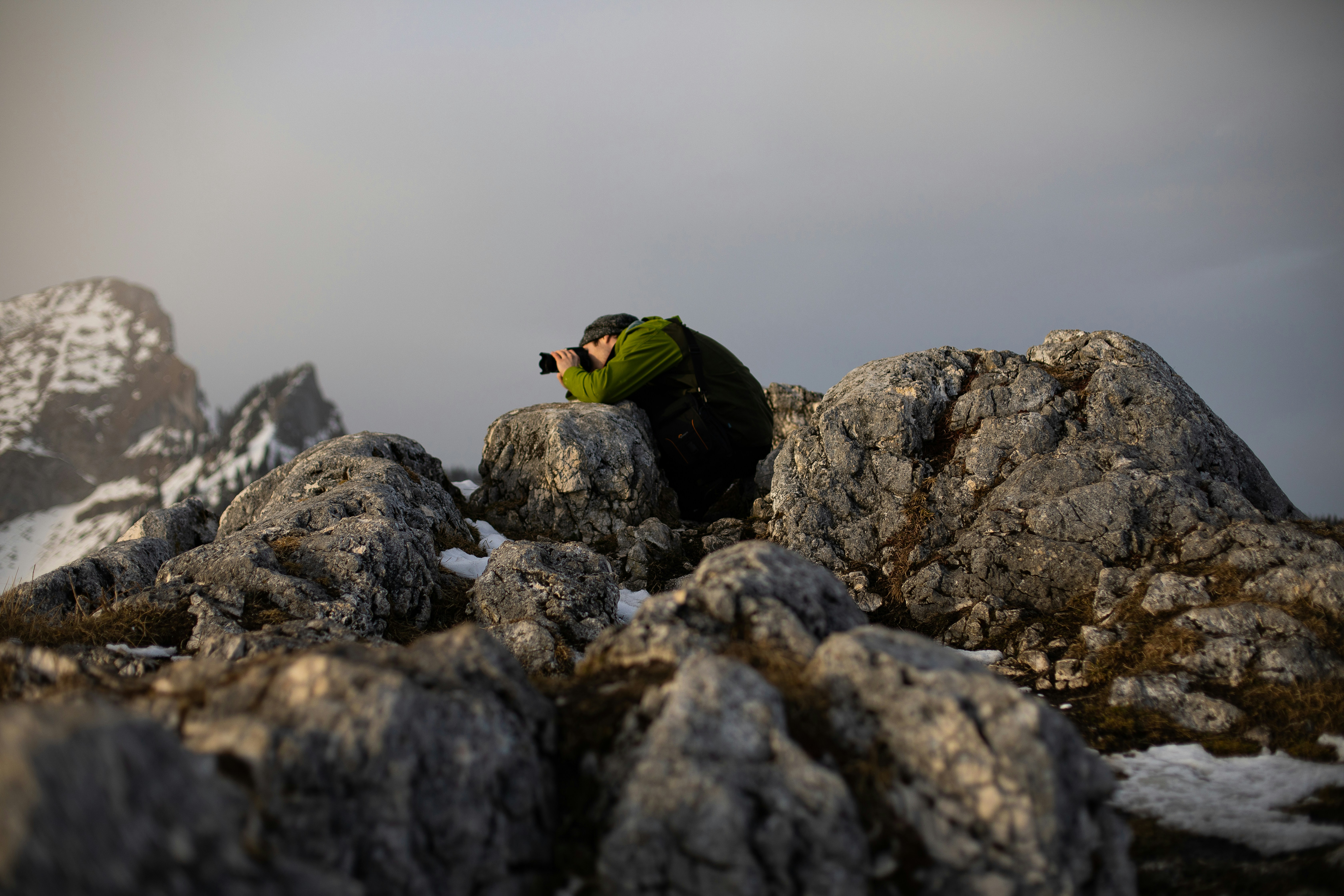 Photographer crouched on rocky terrain, focused on capturing a scene in the mountainous landscape. The rugged environment contrasts with the delicate act of photography.