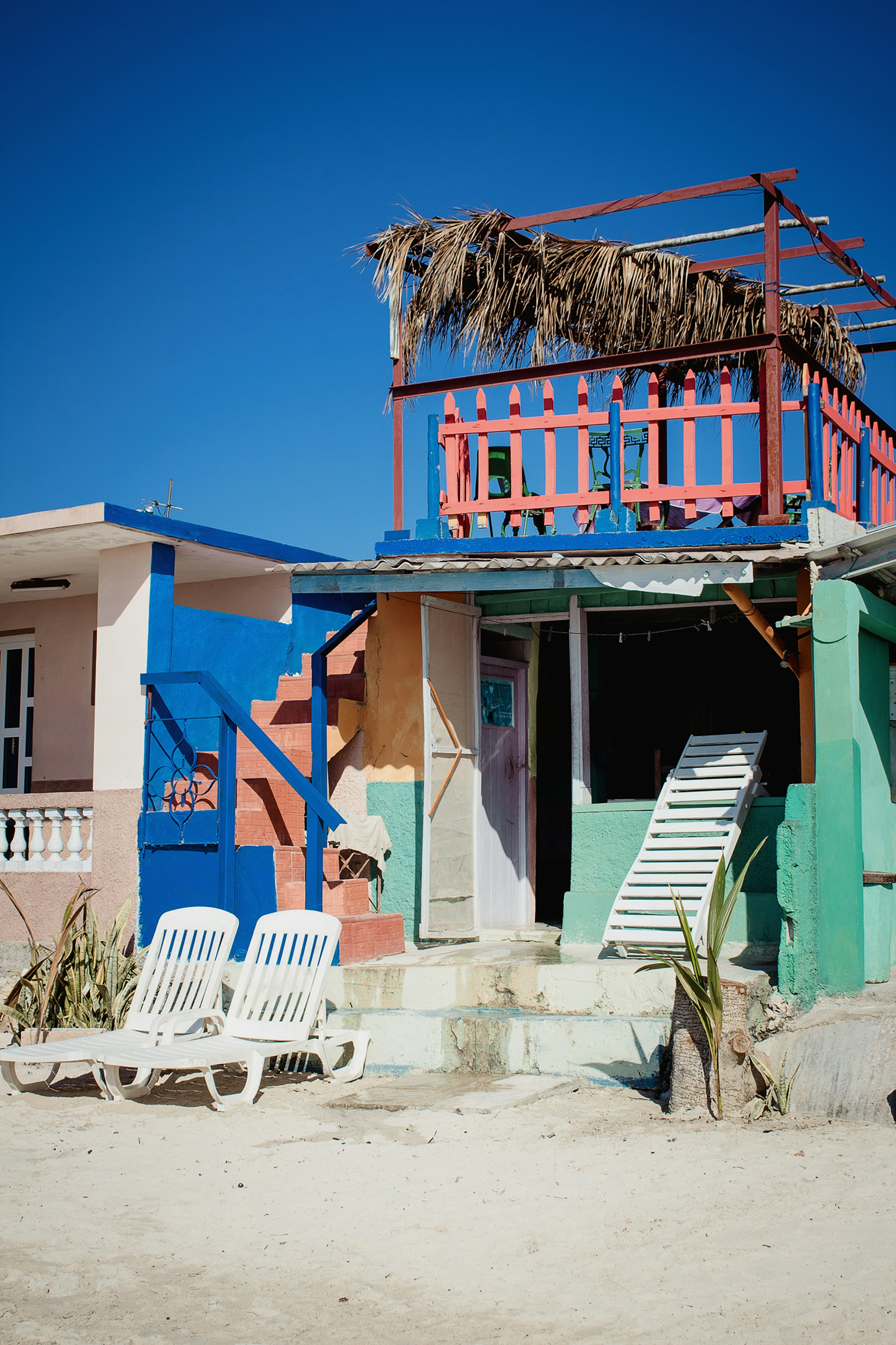 A lively beach house adorned with bright colors and a thatched roof, complemented by white lounge chairs in the foreground. Ideal for a relaxing seaside getaway.