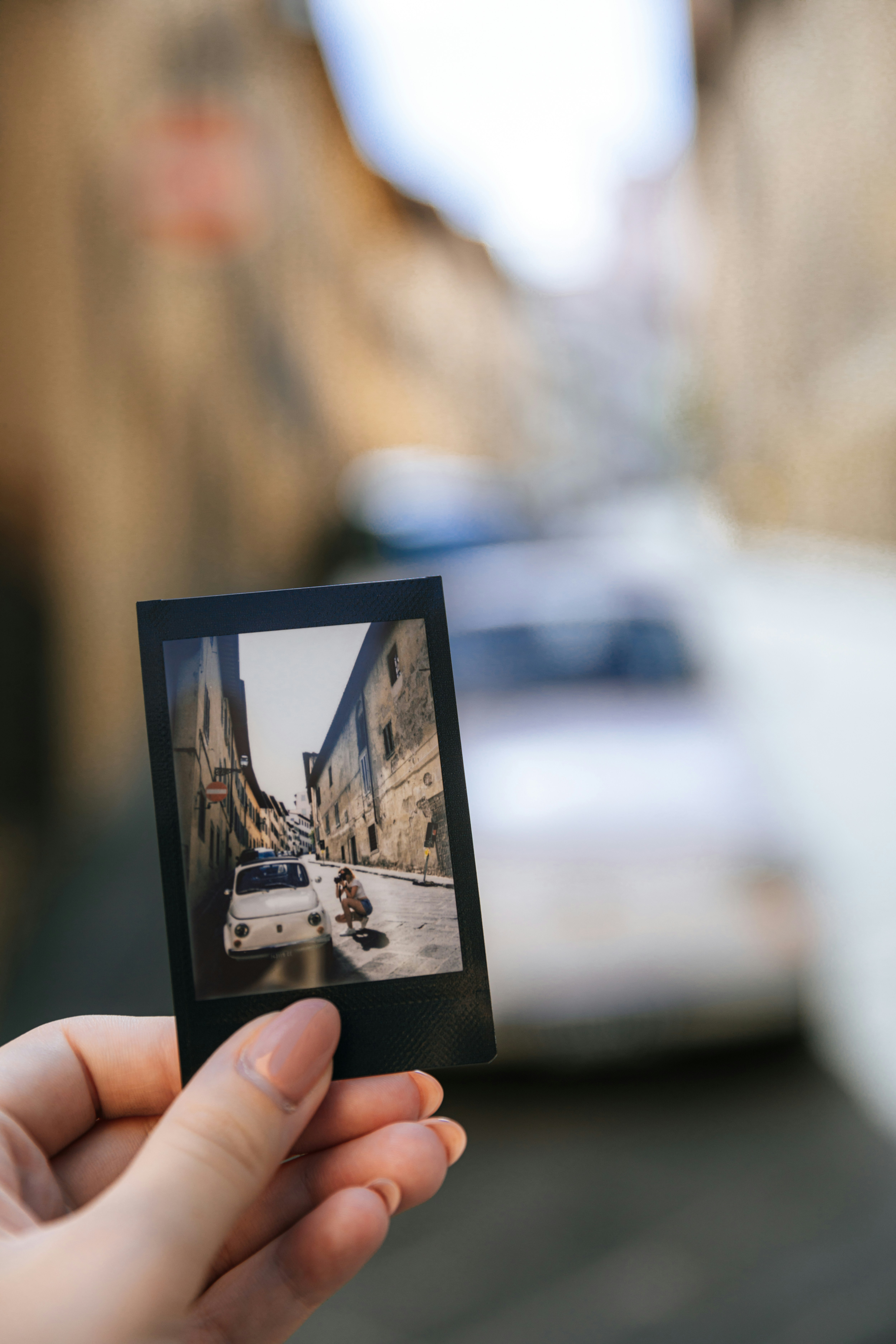 Hand holding a vintage photo of a white car parked on a narrow street, with blurred modern surroundings in the background.