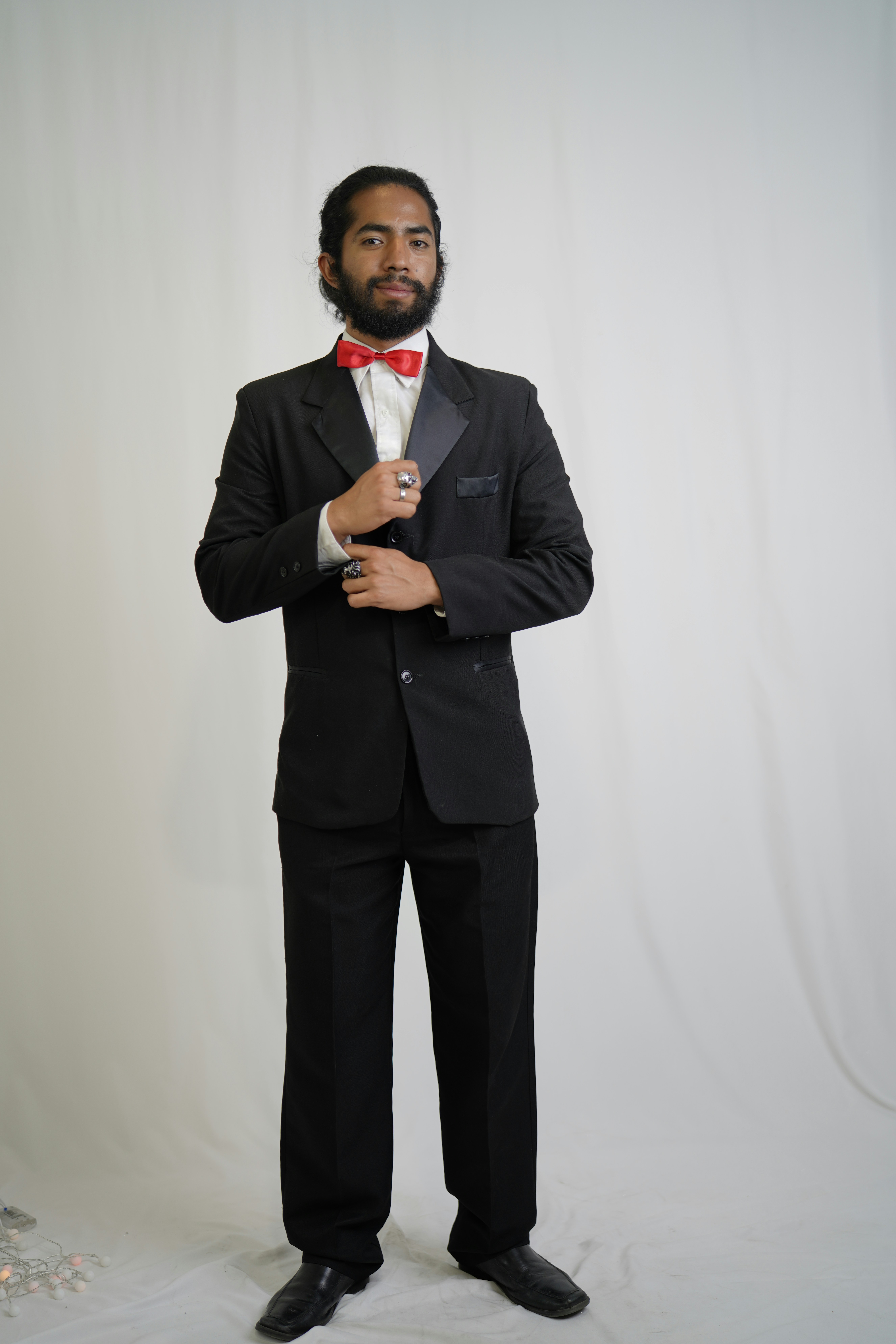Man in a tailored black suit with a red bow tie, striking a confident pose against a neutral backdrop.