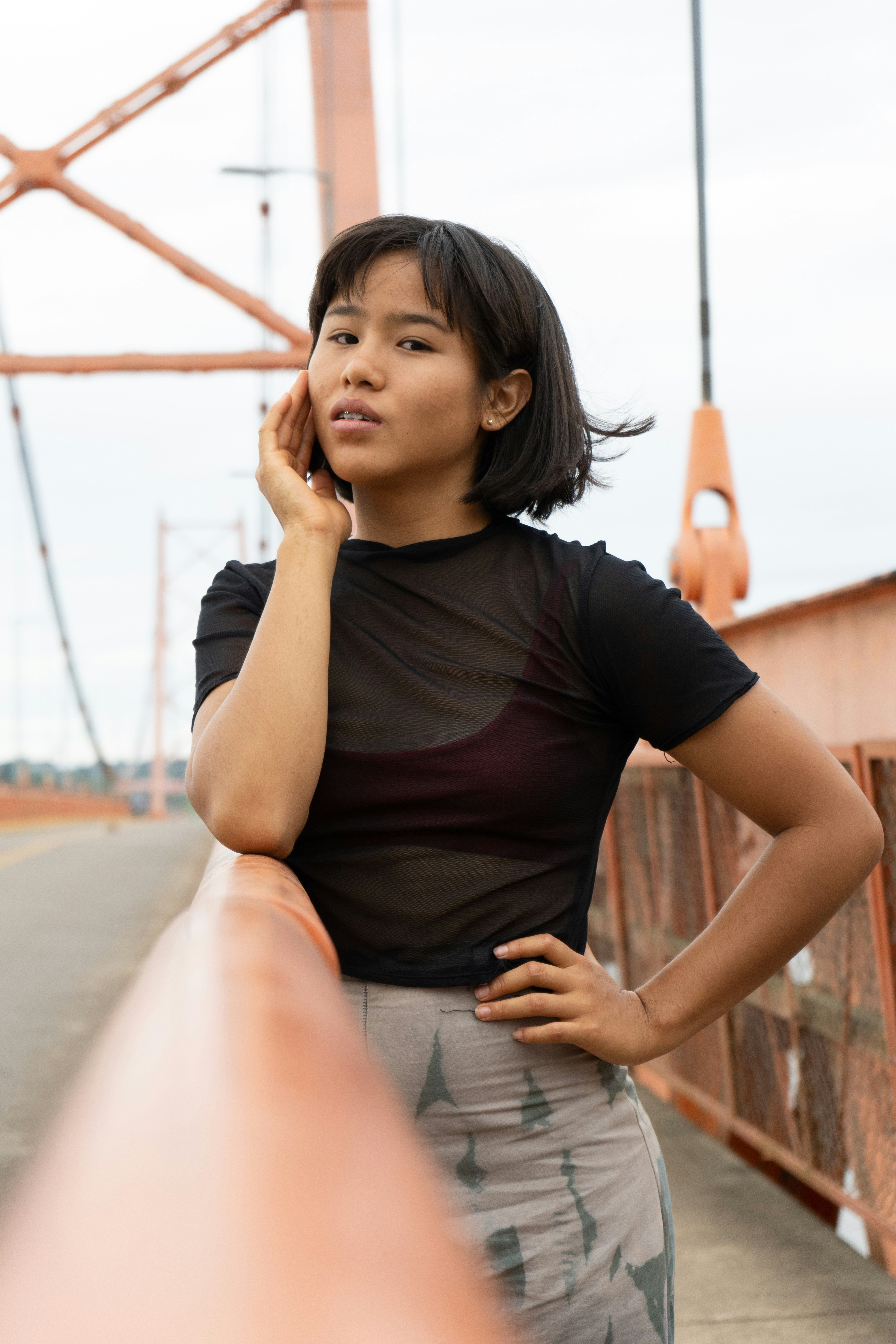 Woman in a black t-shirt and gray pants leans against an orange bridge railing during daytime.