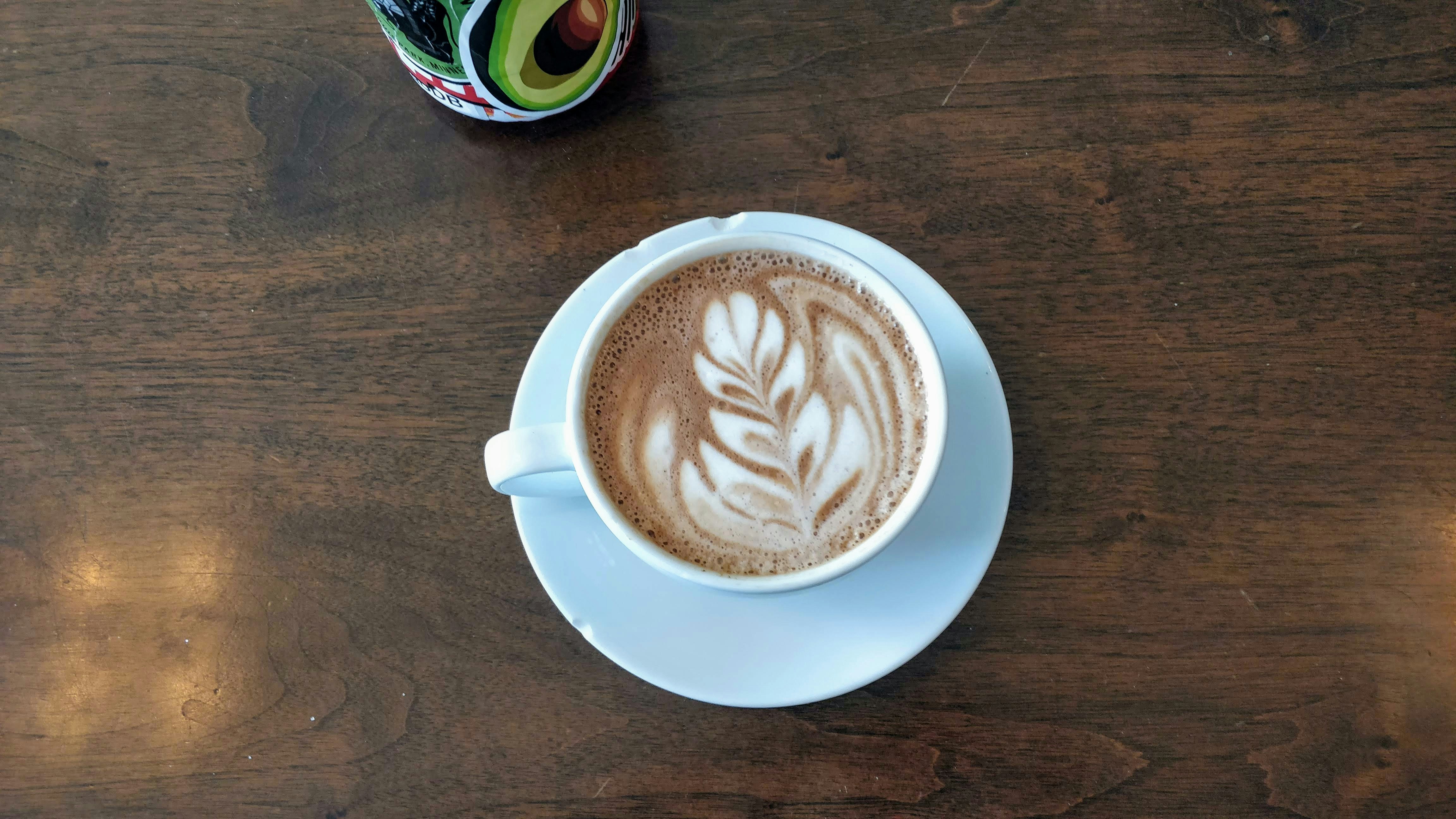 Latte art featuring a delicate leaf pattern sits atop a white cup on a wooden table, accompanied by a colorful beverage in the background.