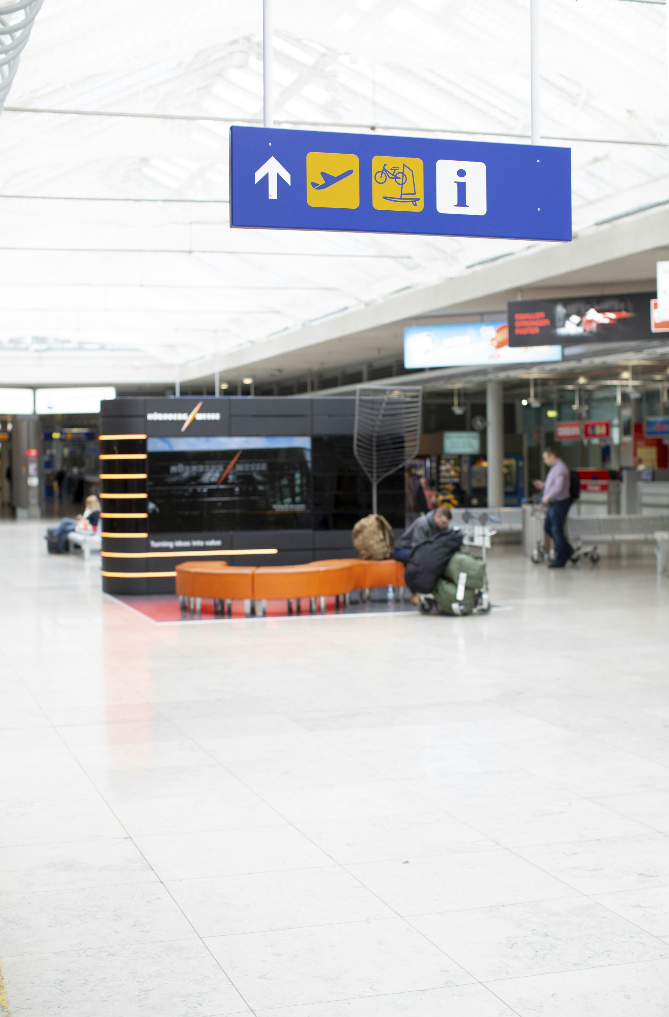 A busy airport terminal showcasing a digital information display alongside bright seating areas. Travelers are seen engaging with their surroundings.