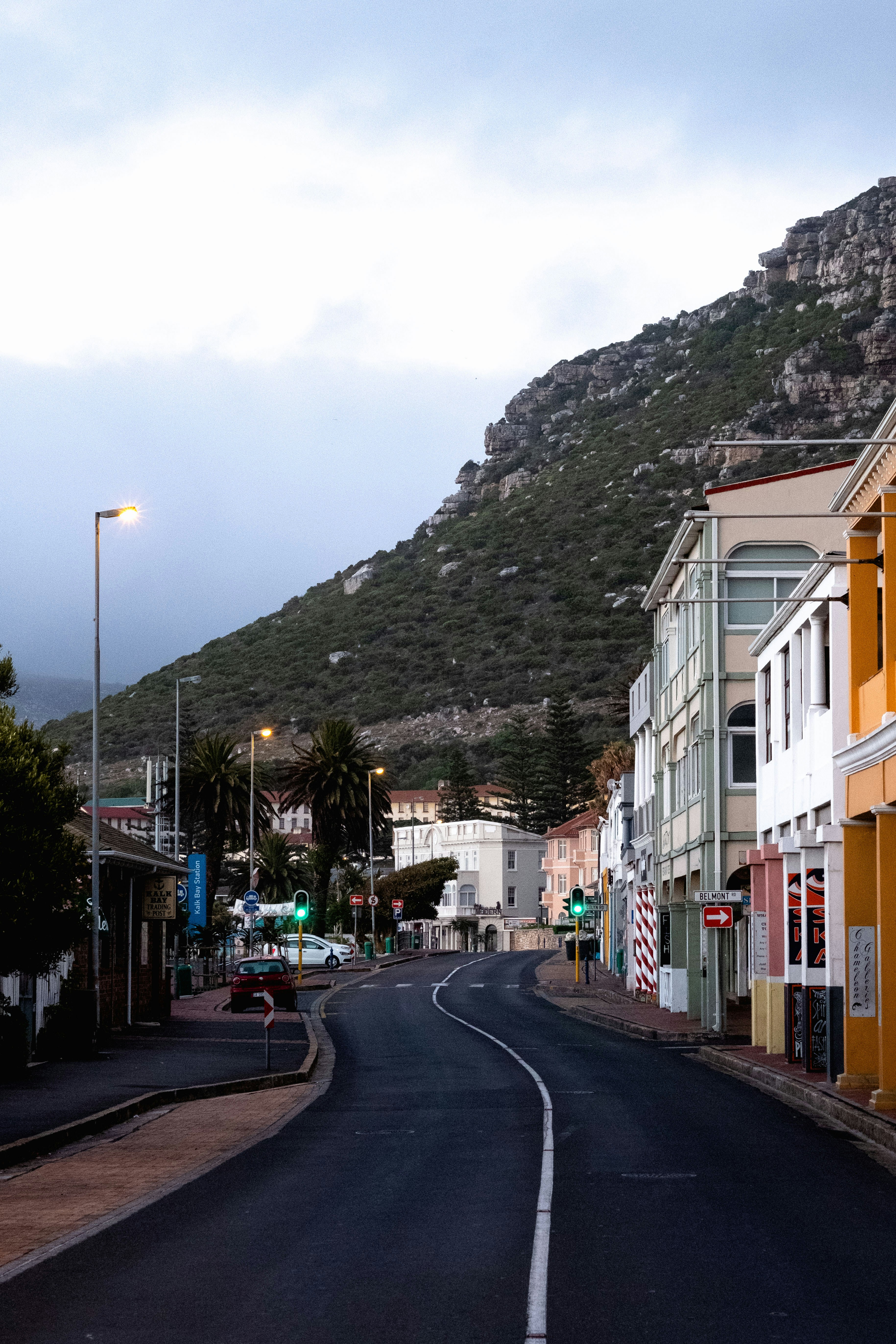 Quiet street lined with colorful buildings and palm trees, leading towards a mountainous backdrop under a cloudy sky.