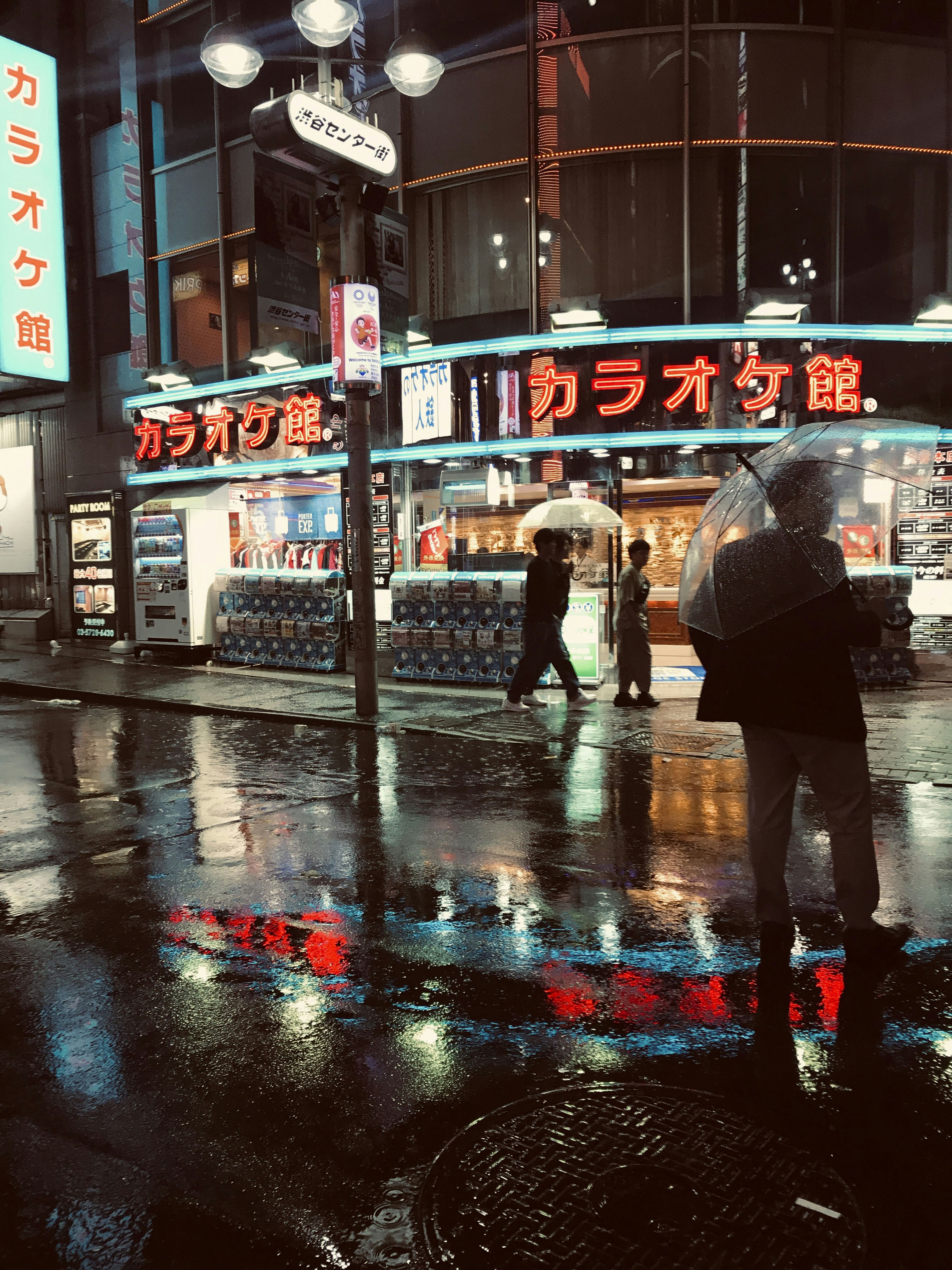 Urban scene featuring pedestrians with umbrellas walking past a karaoke venue illuminated by vibrant neon signs. The wet pavement reflects the colorful lights, enhancing the atmosphere.