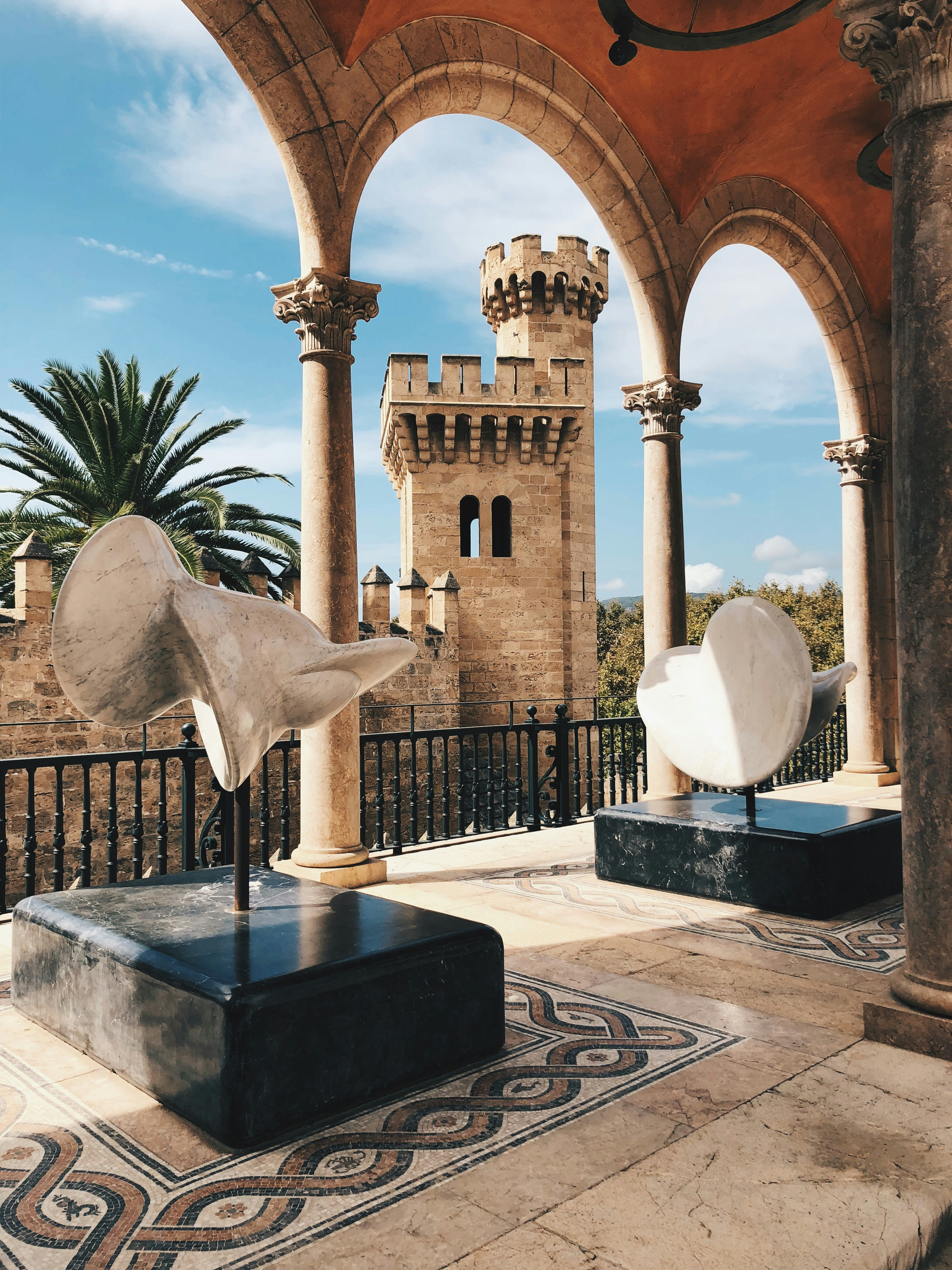 Two modern sculptures stand on pedestals in an ornate archway, framed by a historic tower and lush palm trees in the background.