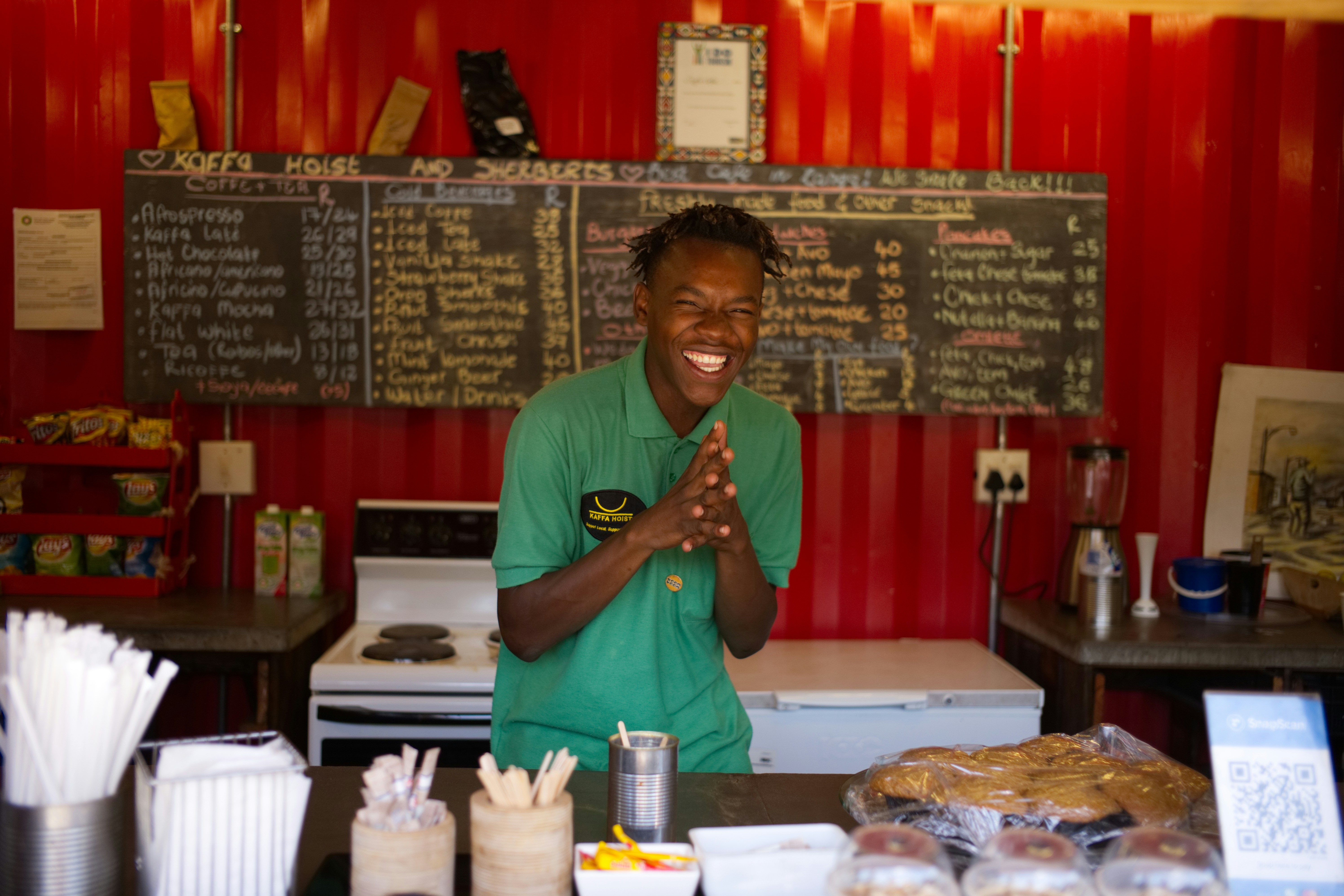 Candid photo of a smiling barista in a green polo behind a counter, with a chalkboard menu and red-walled interior.