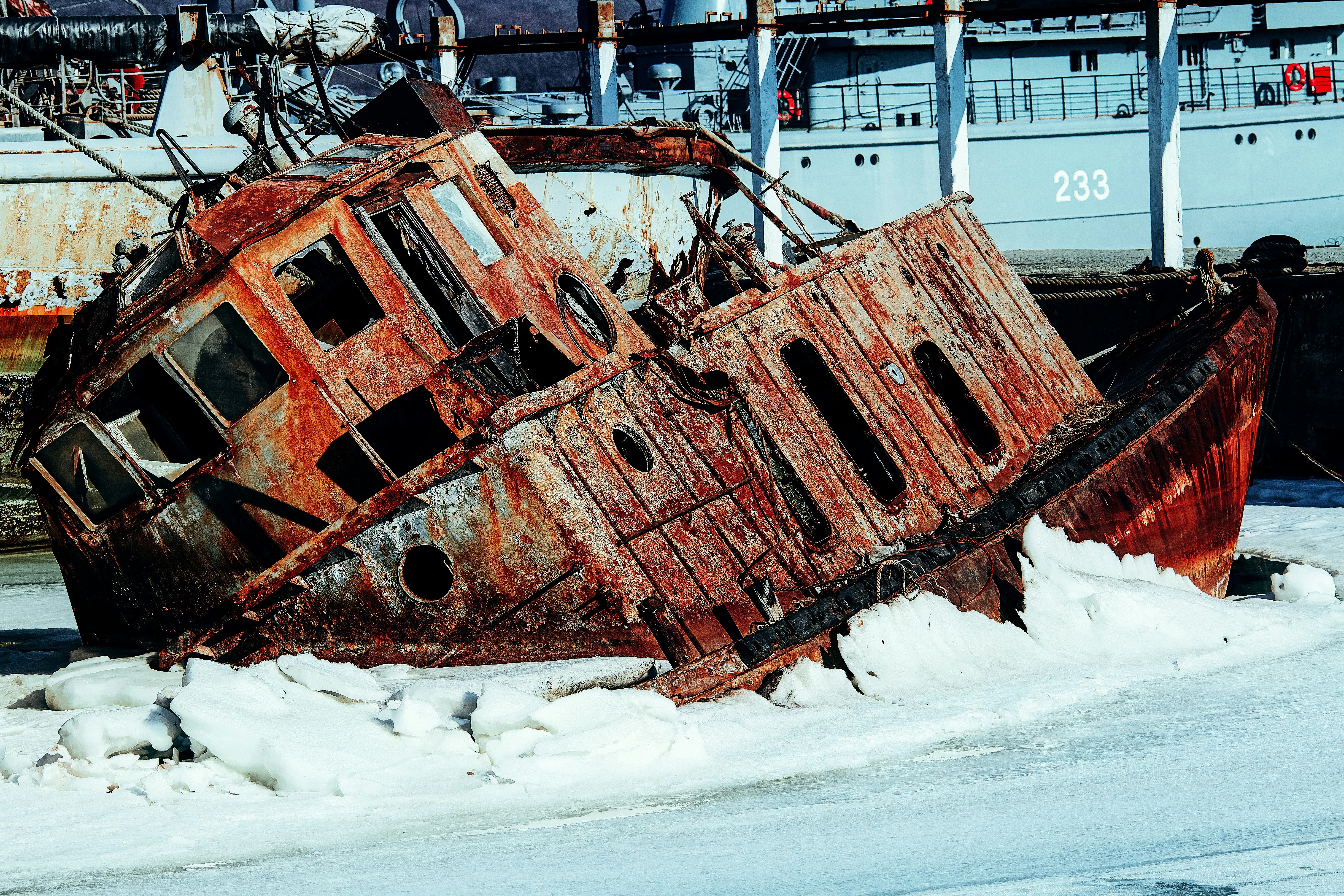 Decaying shipwreck partially submerged in icy waters, showcasing rust and weathered textures against a backdrop of distant vessels.