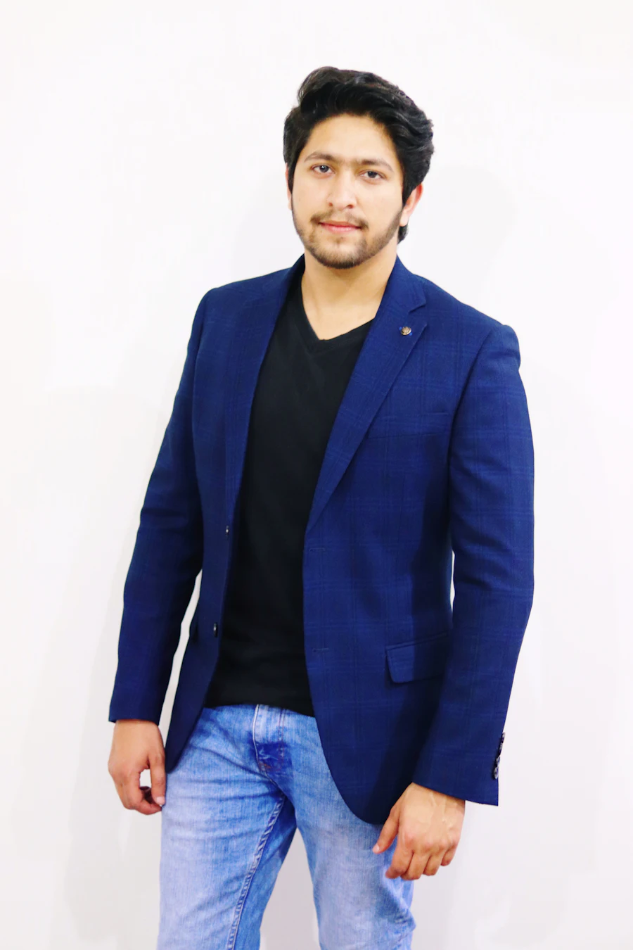 Young man standing against a plain white background, wearing a blue blazer, black shirt, and light jeans. He faces the camera with a relaxed expression.
