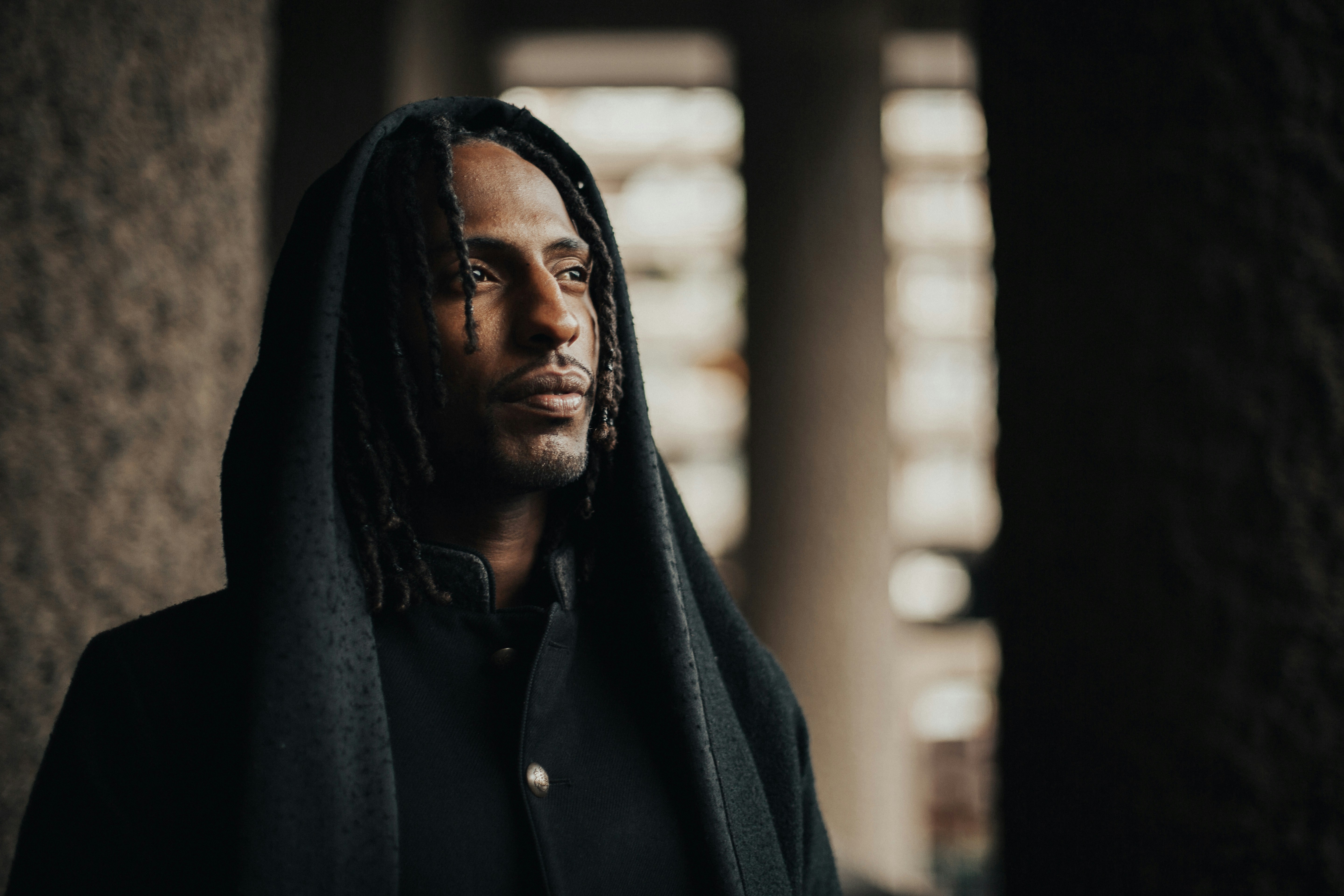 Man with dreadlocks wearing a hooded coat gazes thoughtfully in a dimly lit urban setting, framed by textured pillars.