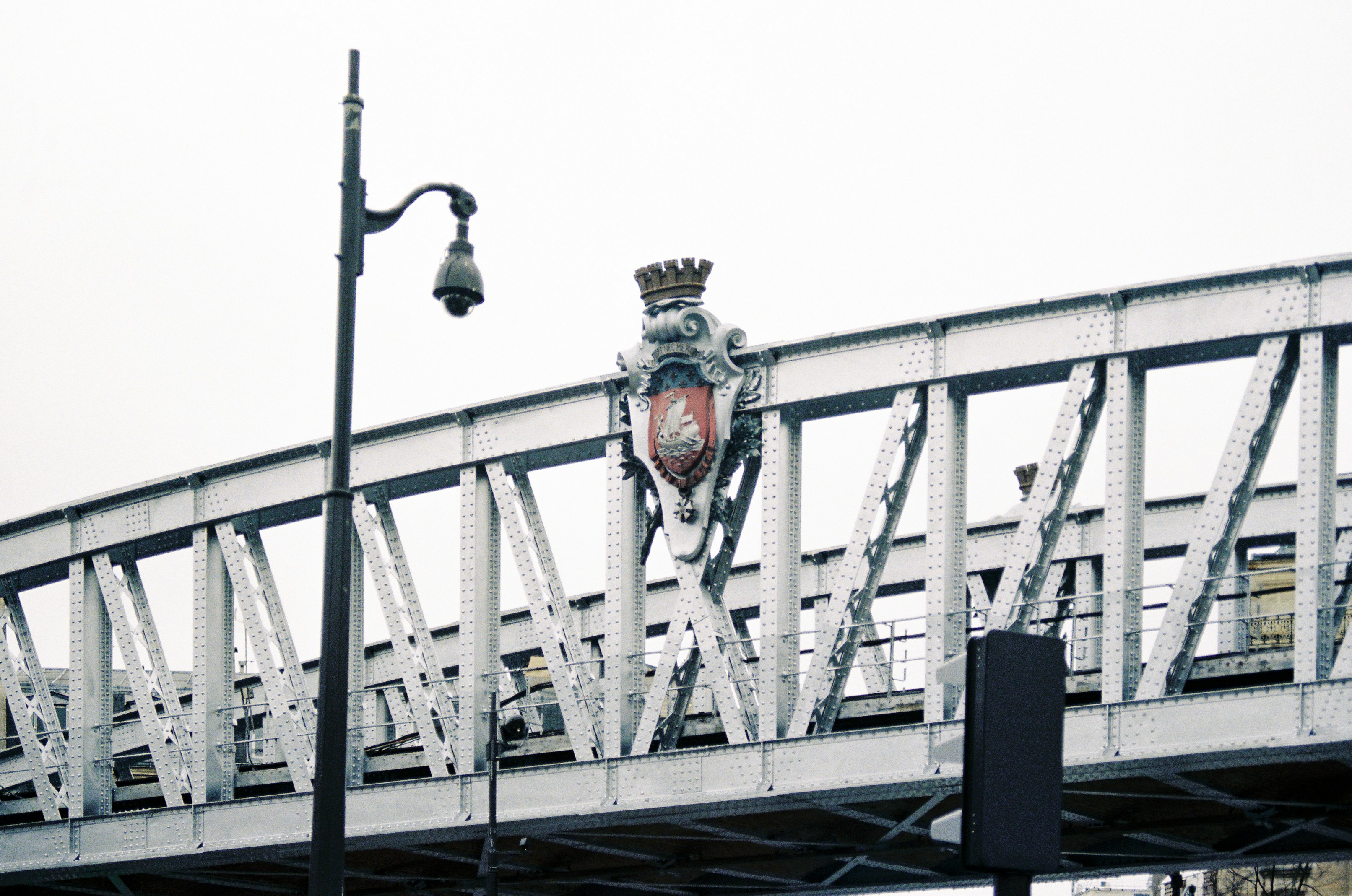 A prominent coat of arms displayed on a metal bridge, showcasing intricate design elements against a muted sky.
