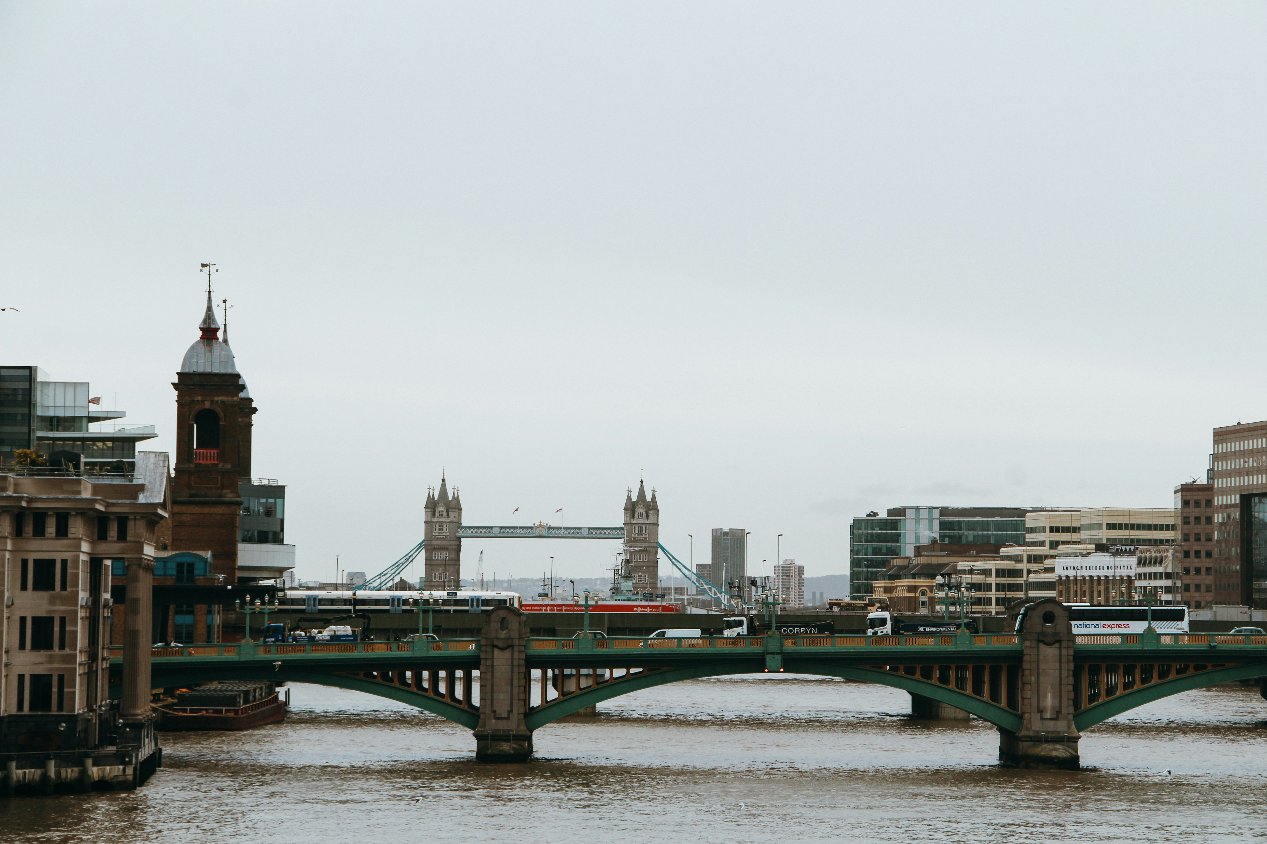 View of Tower Bridge and the Thames River, framed by urban architecture and a historic clock tower.