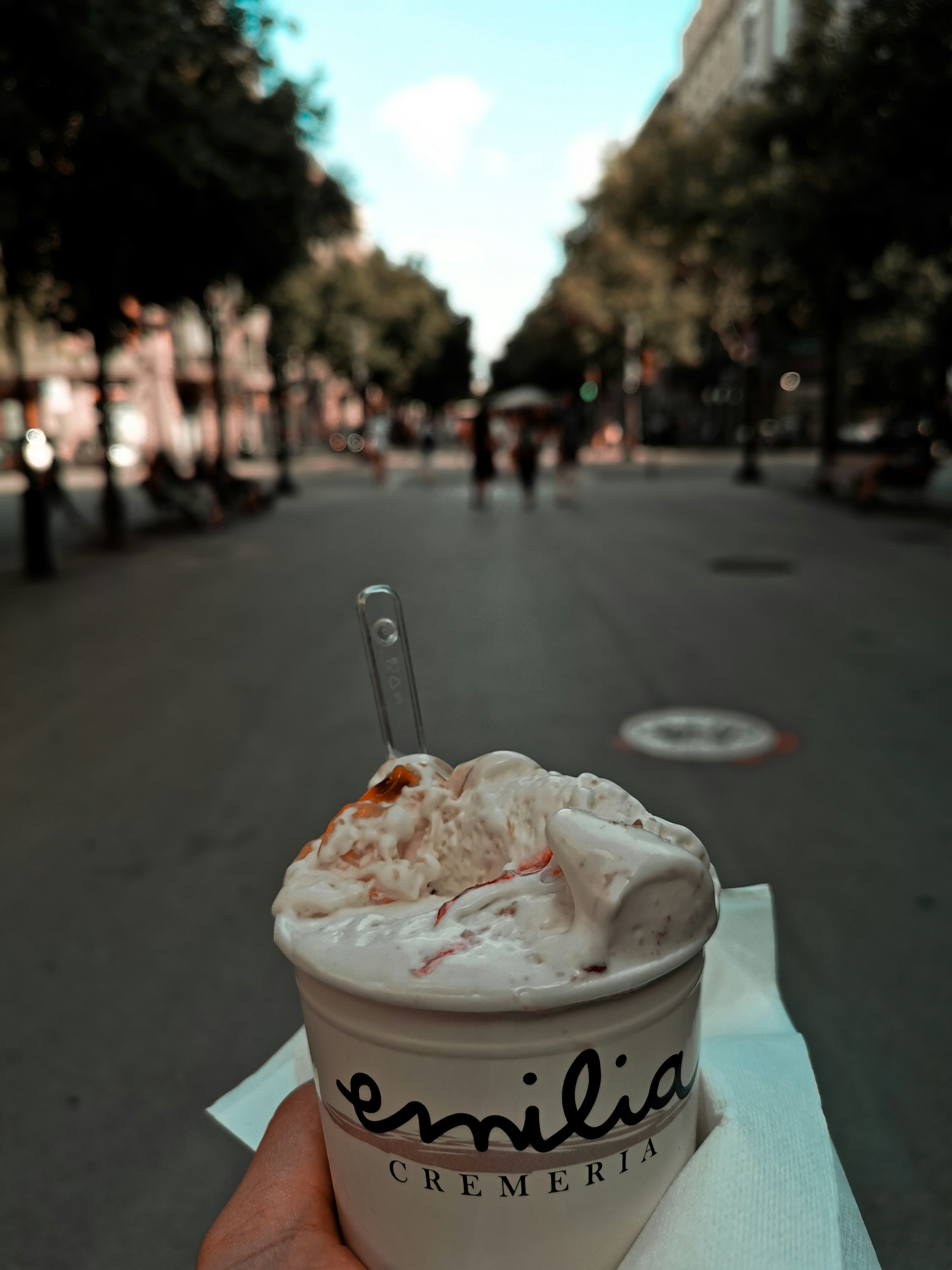 Cup of creamy ice cream held in hand, with a bustling street scene blurred in the background. The logo 'emilia' is prominently displayed on the cup.