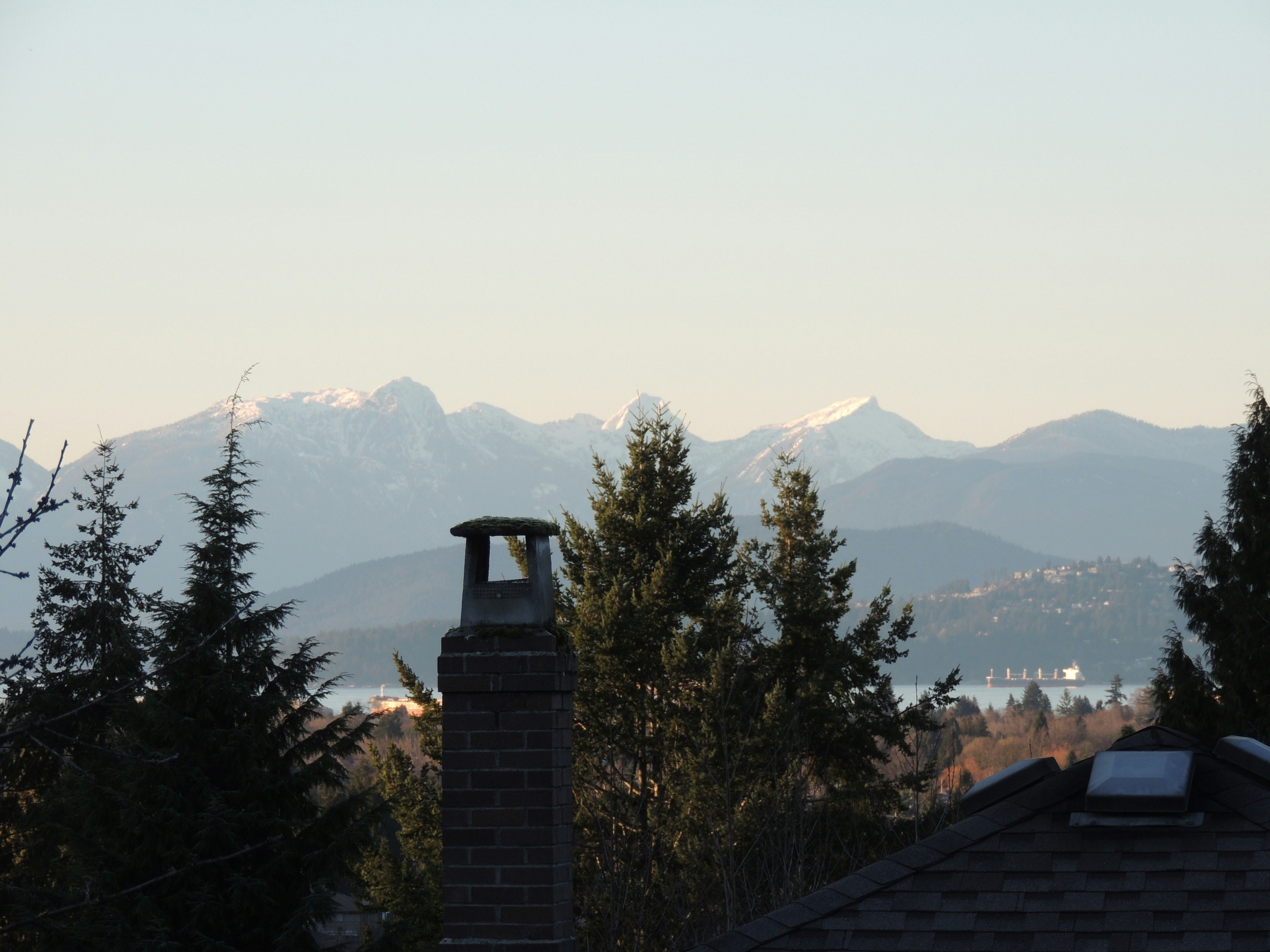 Snow-capped mountains rise majestically behind a silhouette of trees and rooftops, capturing the essence of a tranquil evening. 