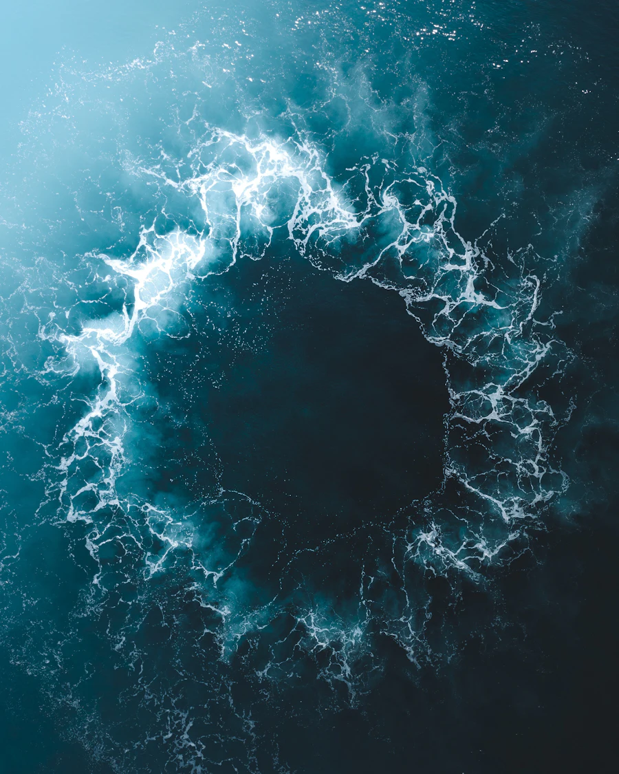 Top-down view of a swirling ocean ring with bright white foam surrounding a dark center. Deep teal water fills the frame.