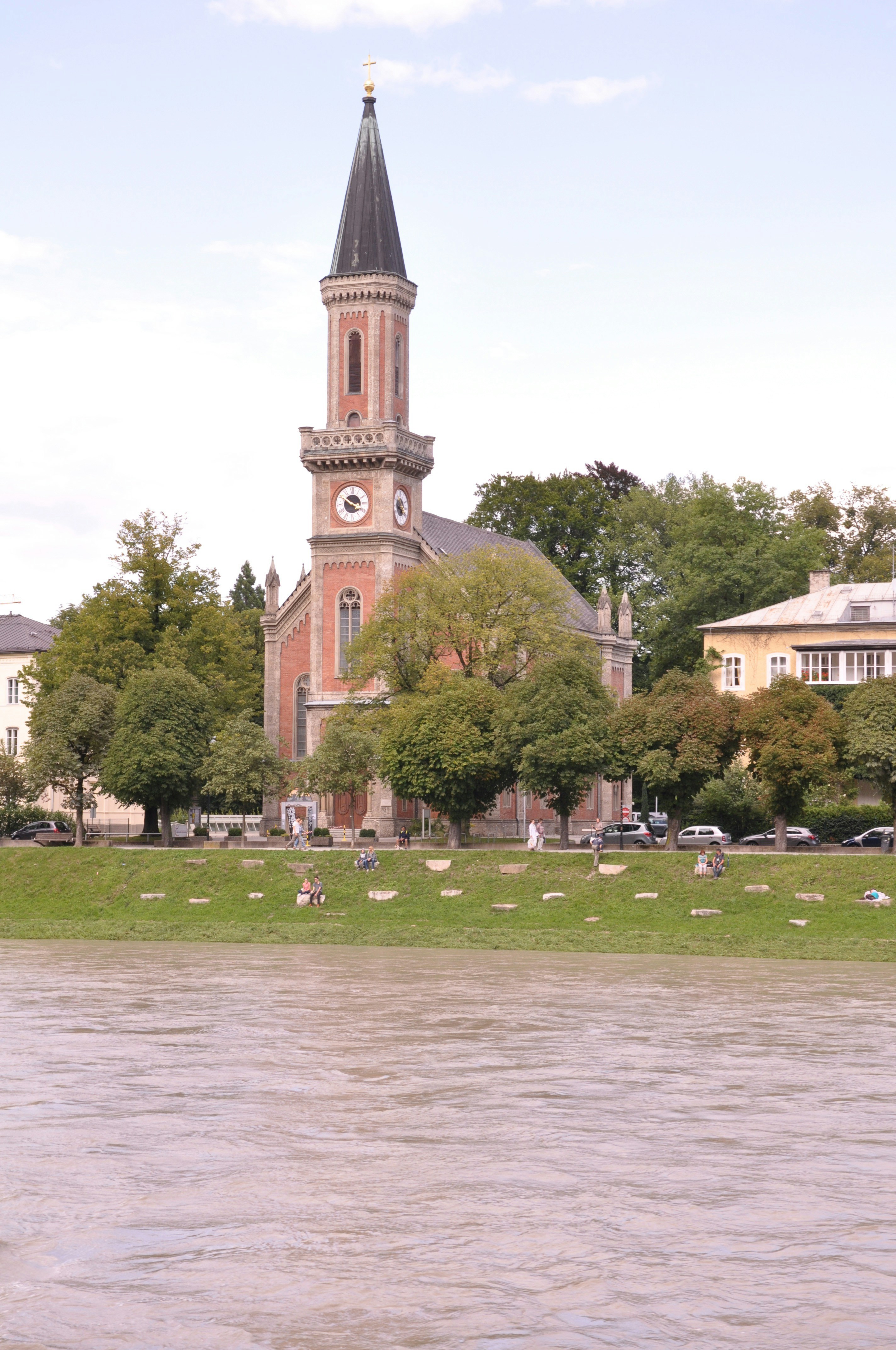 Historic church nestled among lush trees along a riverbank, with people enjoying the serene landscape.