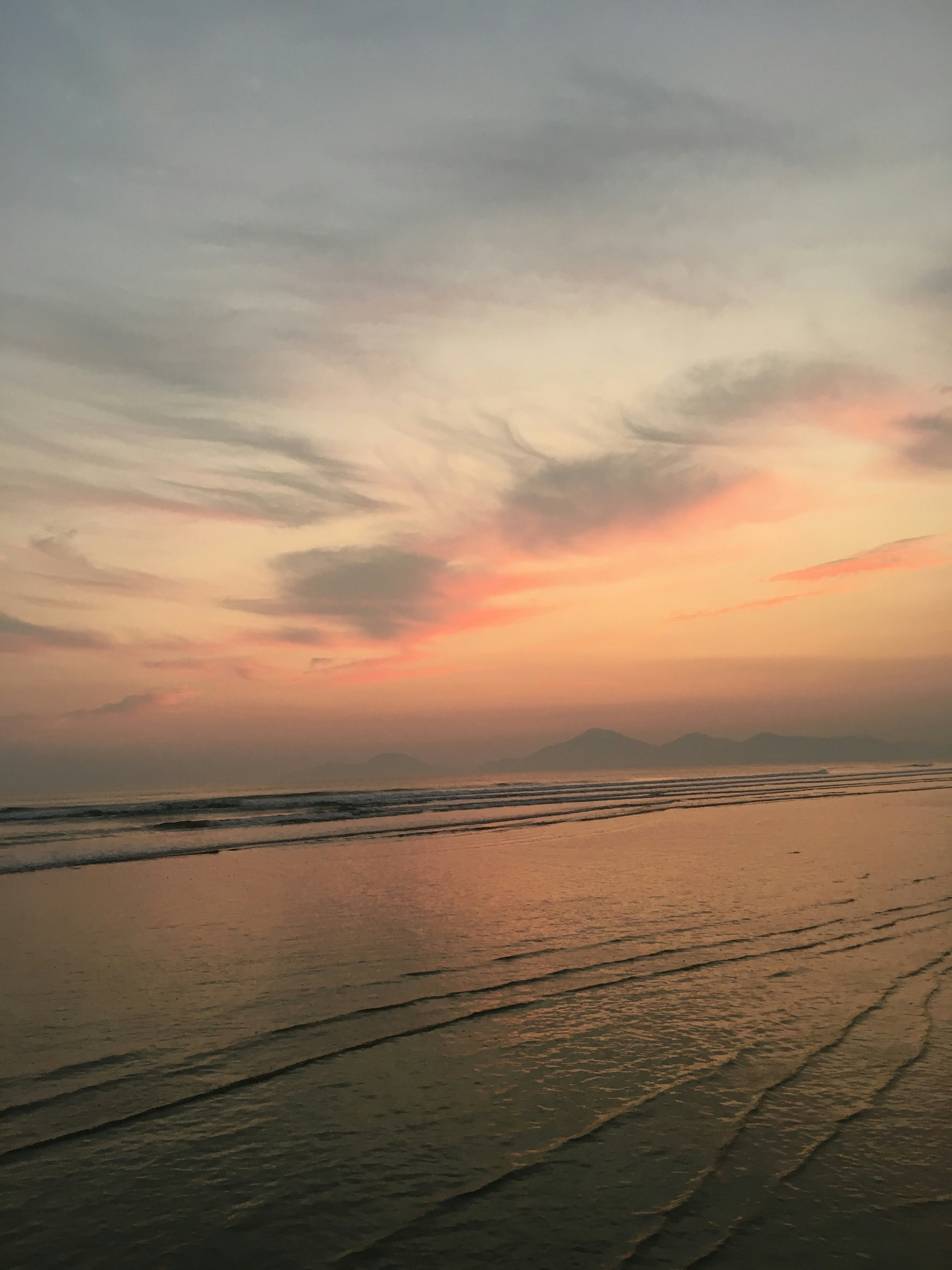 Serene beach scene at sunset, with gentle waves reflecting warm hues of orange and pink in the sky. Mountains loom softly in the distance.