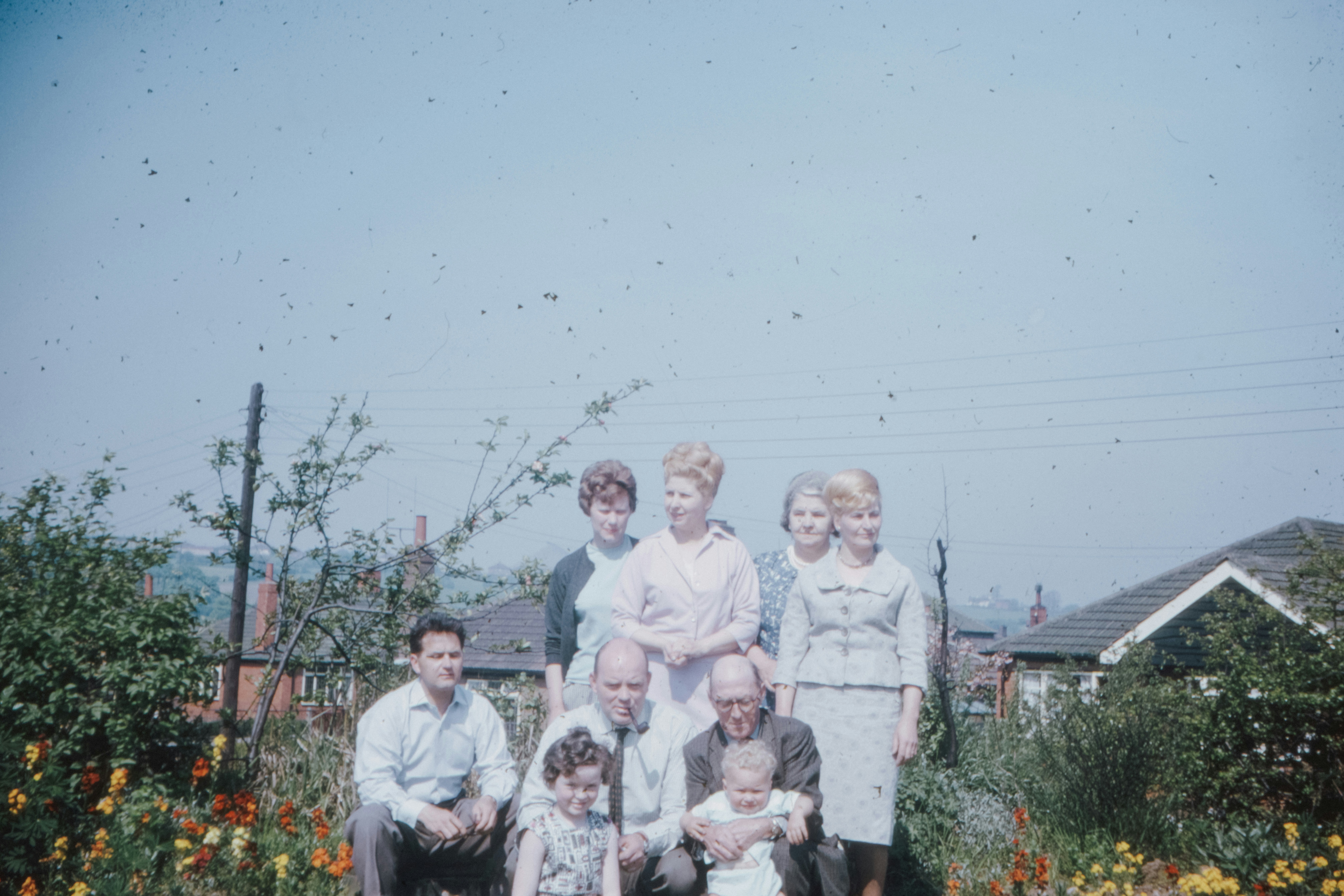 Family gathered in a garden filled with vibrant flowers, capturing a moment of togetherness. The scene reflects a nostalgic atmosphere with a clear blue sky.