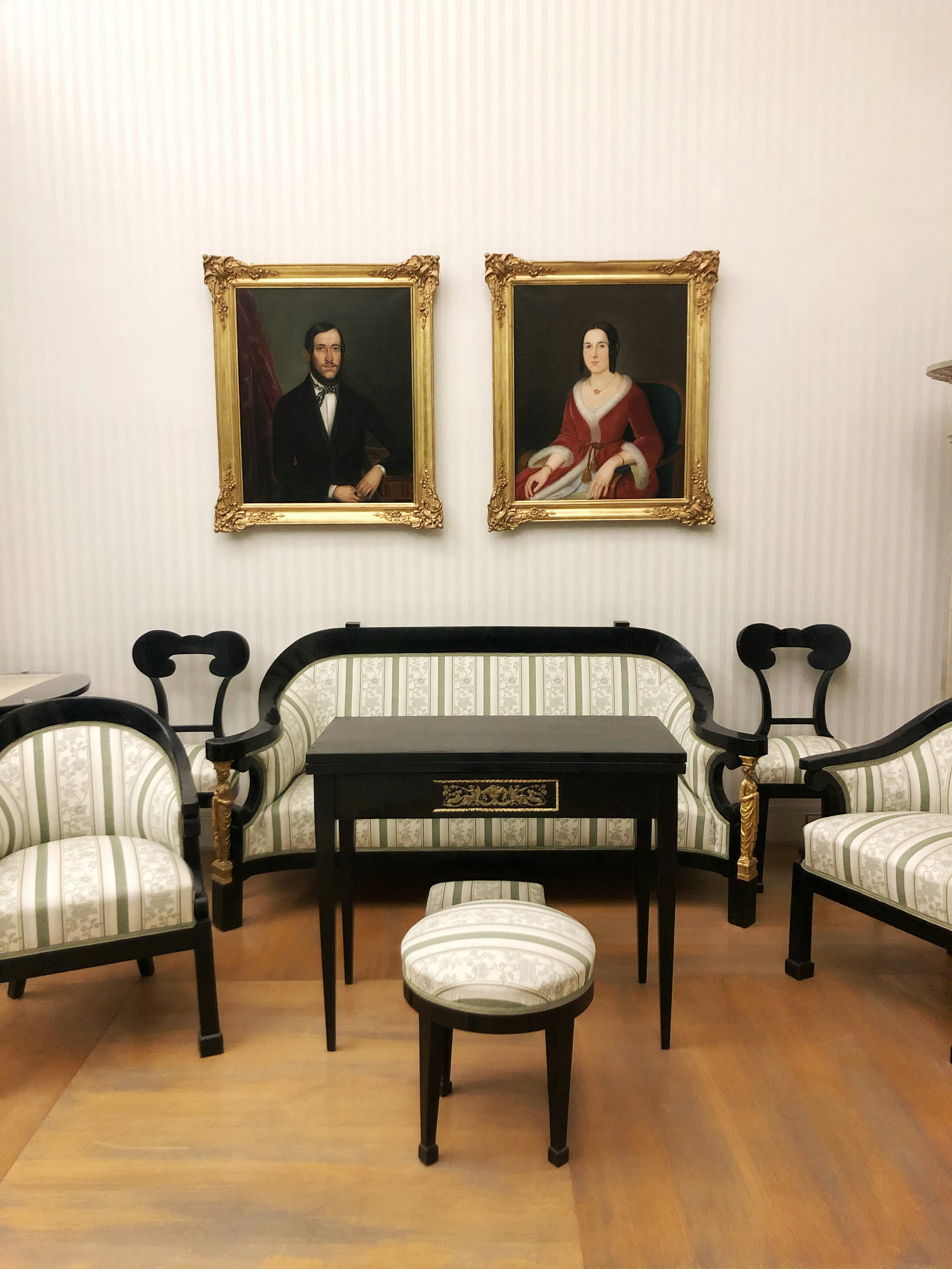 Vintage furniture arrangement featuring a central table with decorative chairs, complemented by two classic portraits on the wall. 