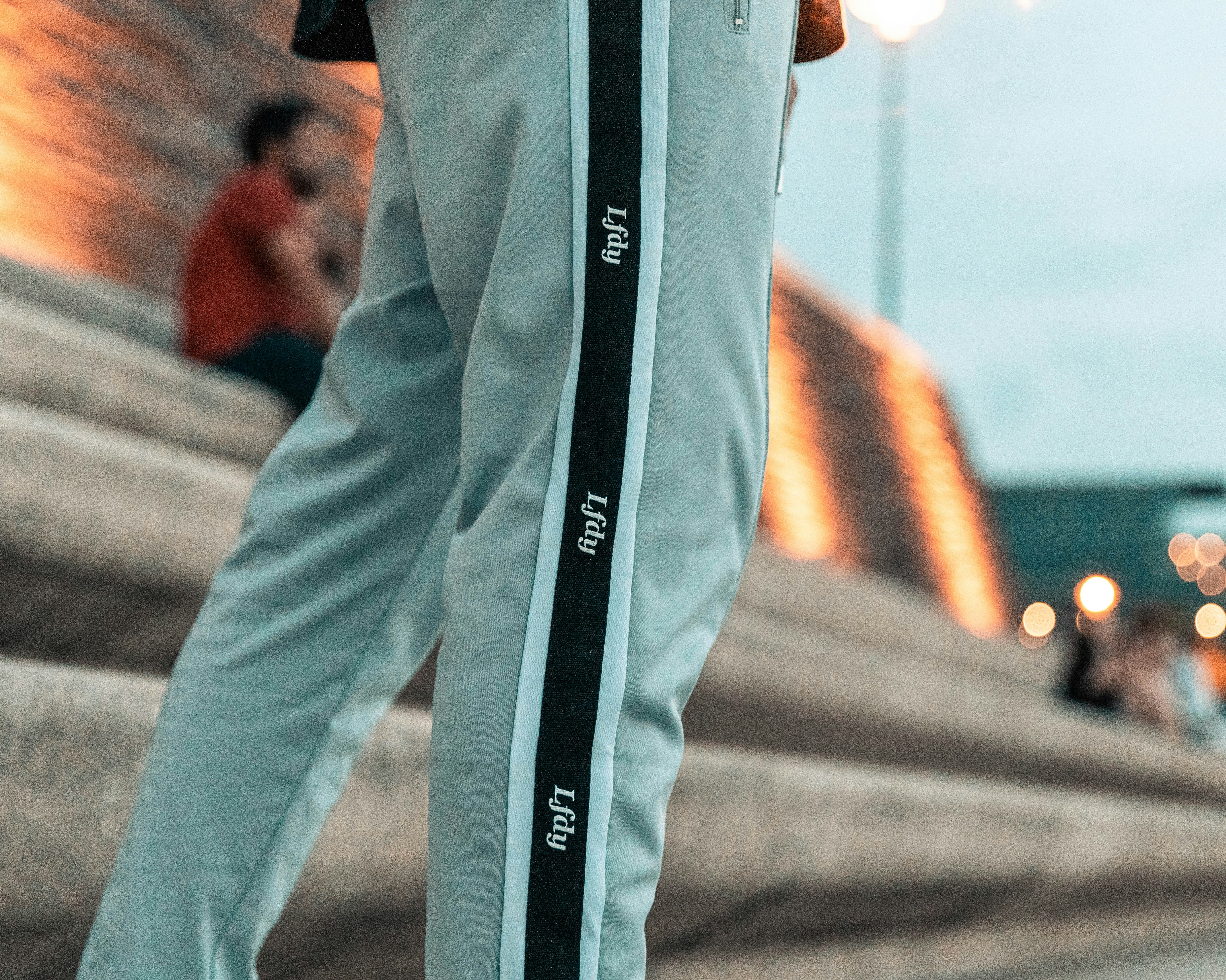 Close-up of gray athletic pants featuring a bold side stripe, set against a blurred urban background with warm lights and figures in the distance.