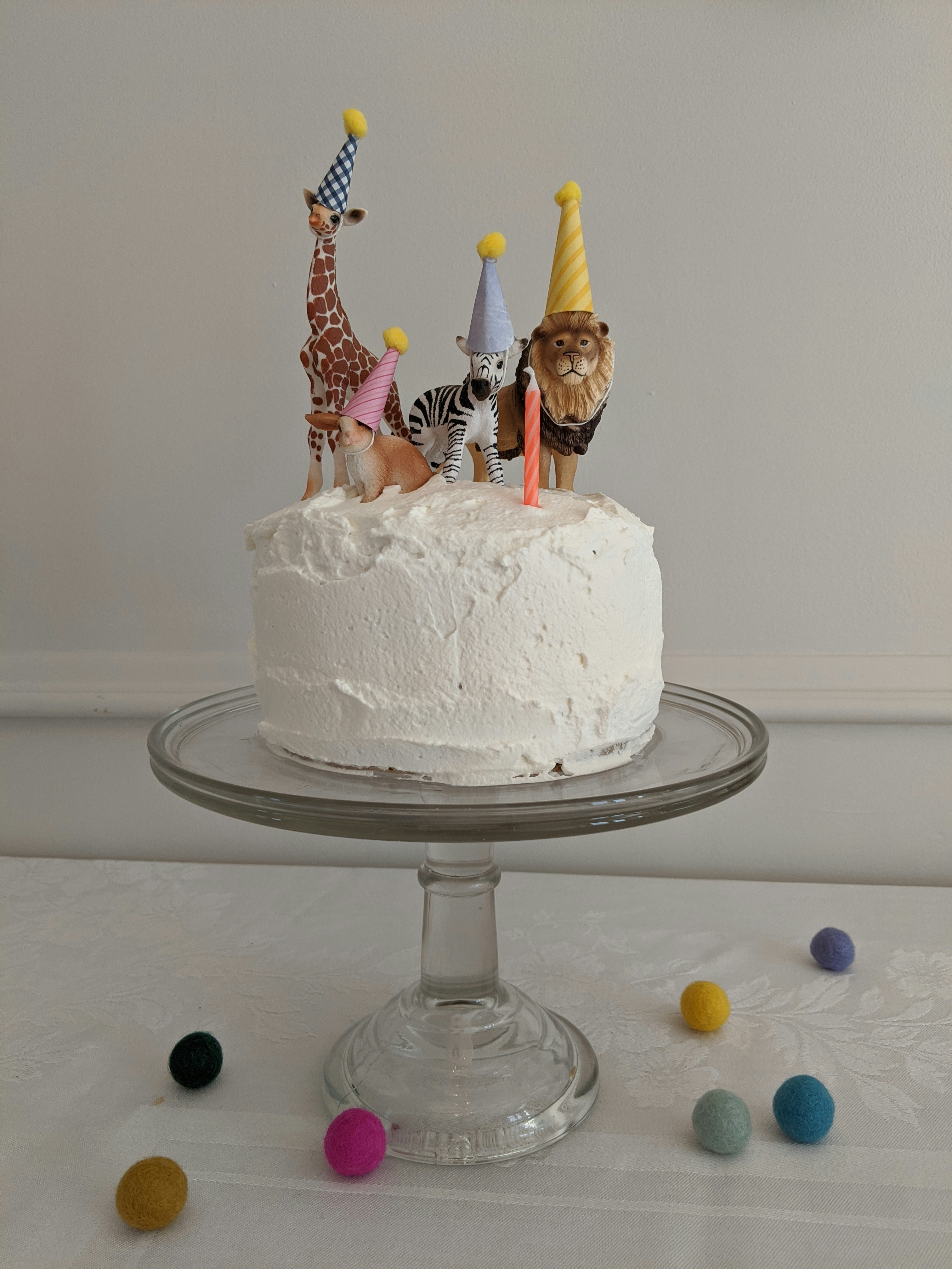 A festive cake topped with playful animal figurines wearing party hats and candles, set on a glass stand with colorful felt balls scattered around.